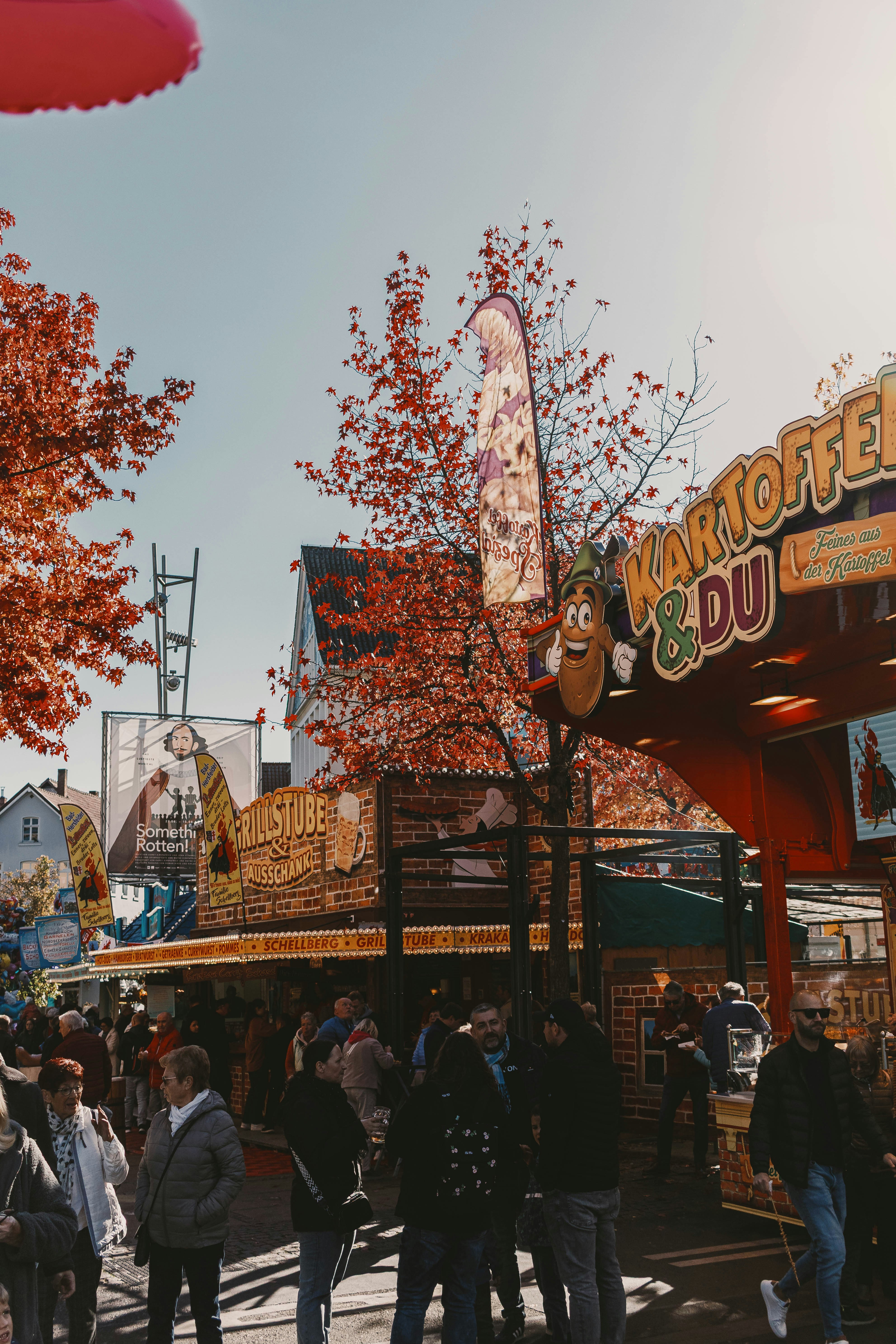 Crowd enjoying a vibrant autumn fair with colorful stalls and trees adorned with red leaves.