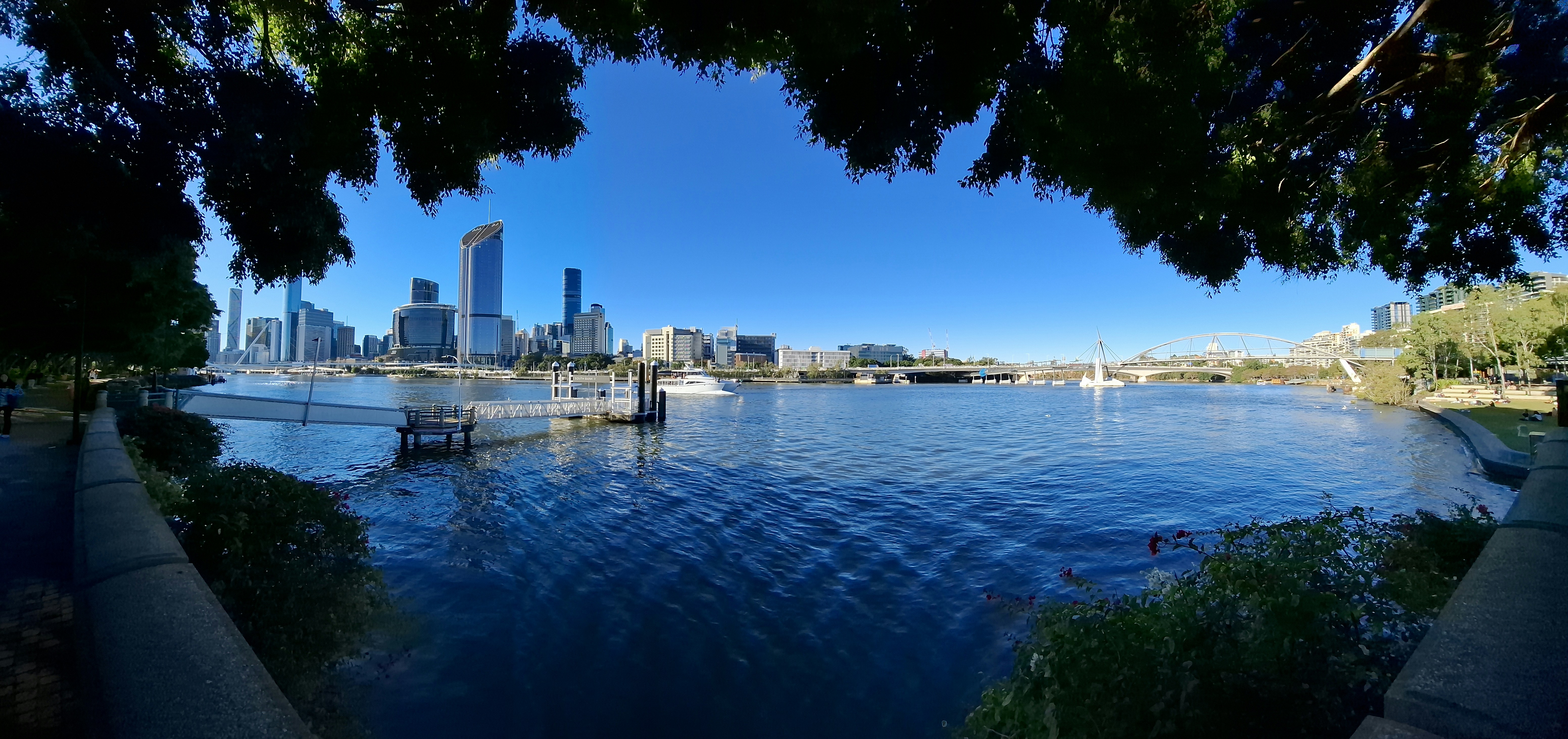 A panoramic view capturing the tranquility of a city waterfront, framed by lush greenery and modern architecture. The scene reflects a perfect blend of nature and urban life.