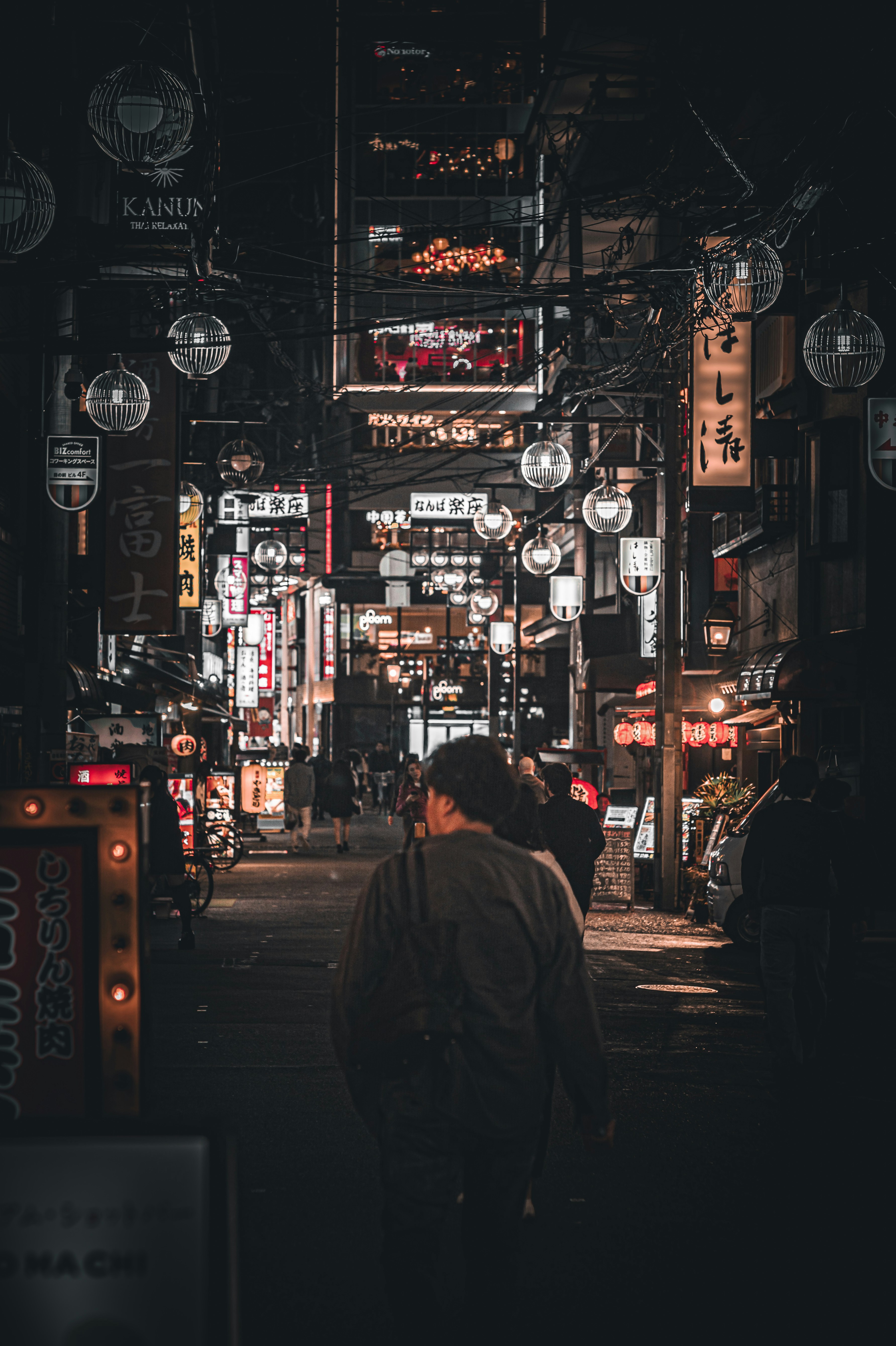 Dimly lit street adorned with vibrant lanterns and neon signs, featuring a solitary figure walking towards the bustling heart of the city.