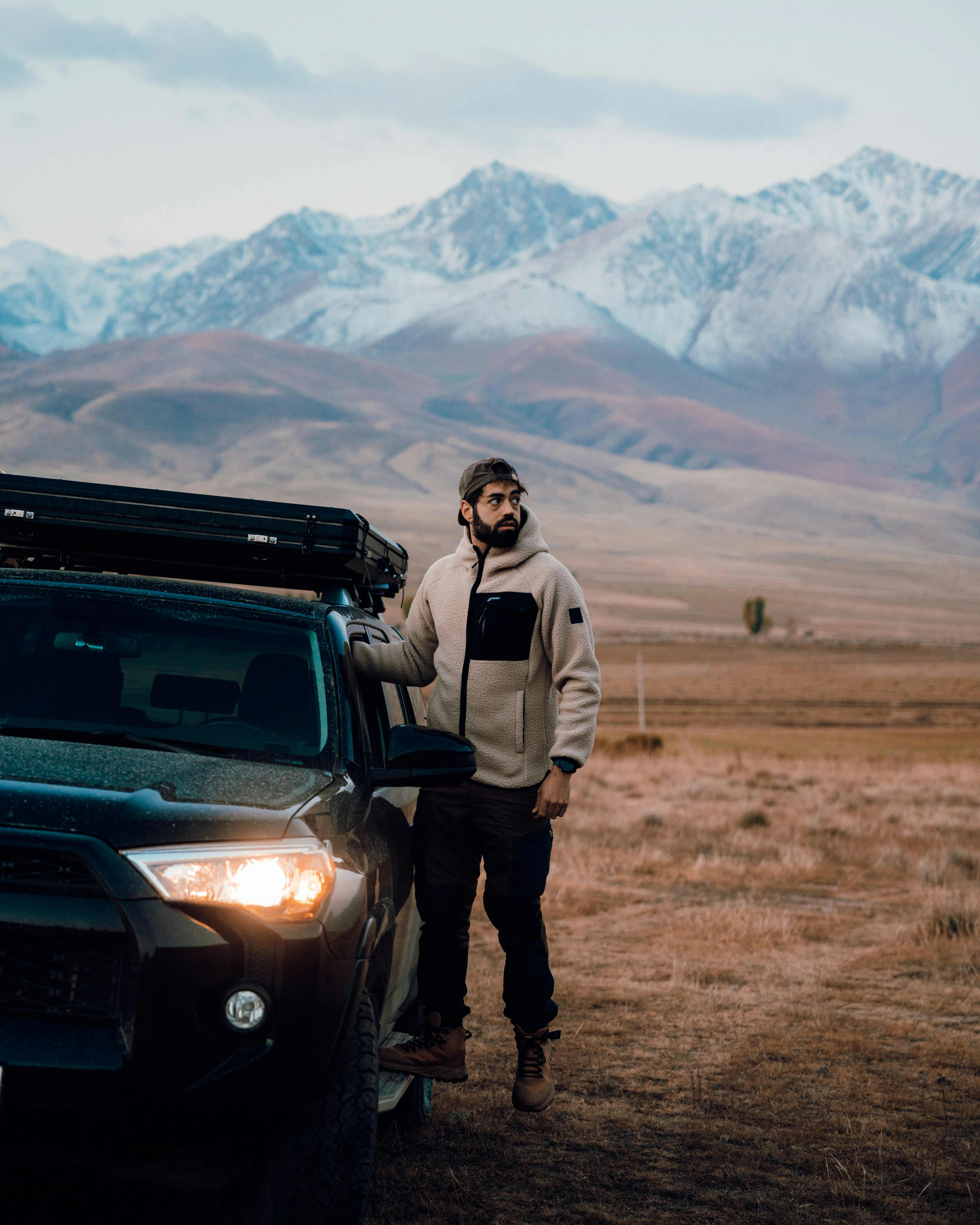 A man stands beside a rugged vehicle, gazing towards distant snow-capped mountains in a serene landscape. The scene captures the essence of exploration and tranquility.