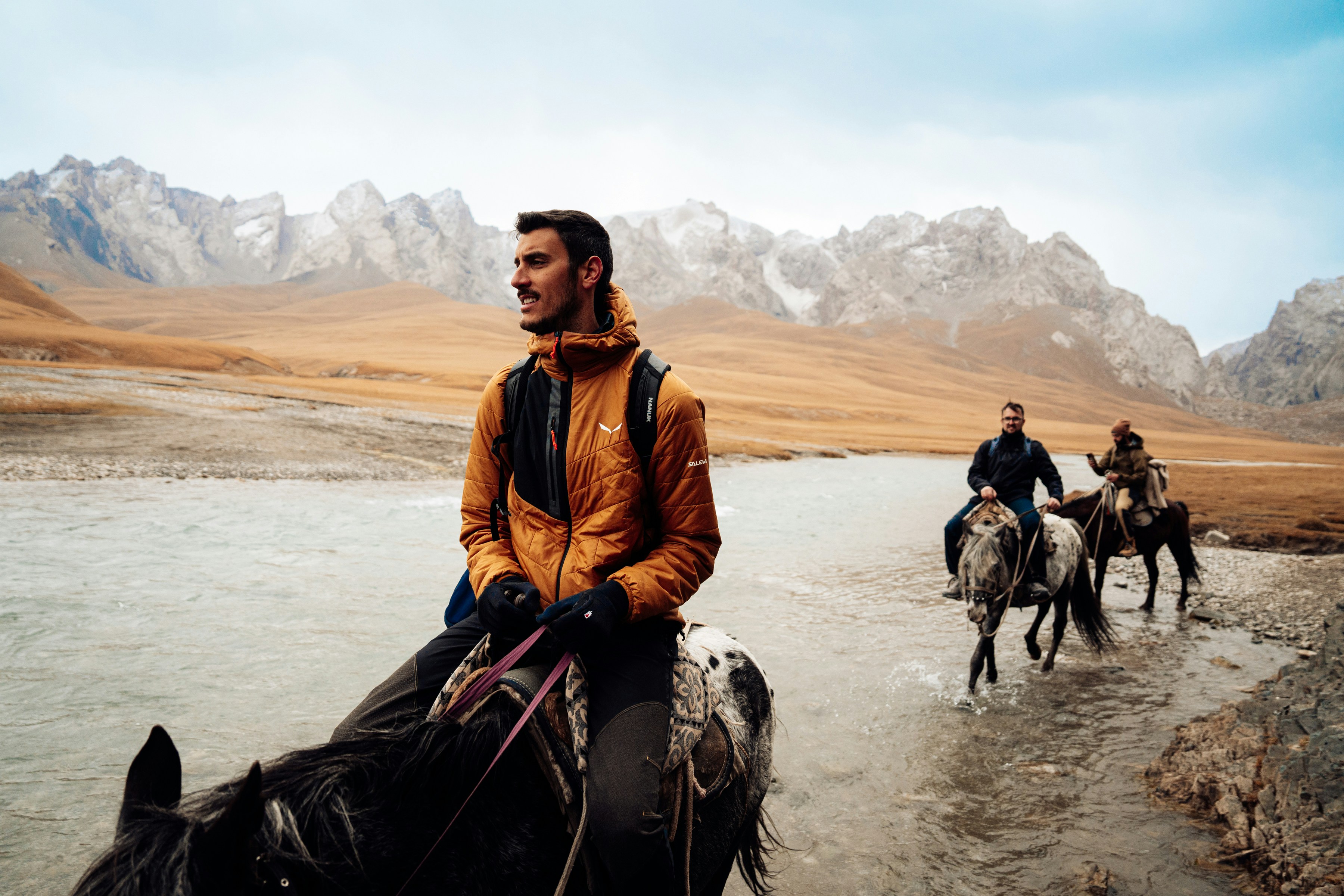 Three people riding horses through a shallow river.