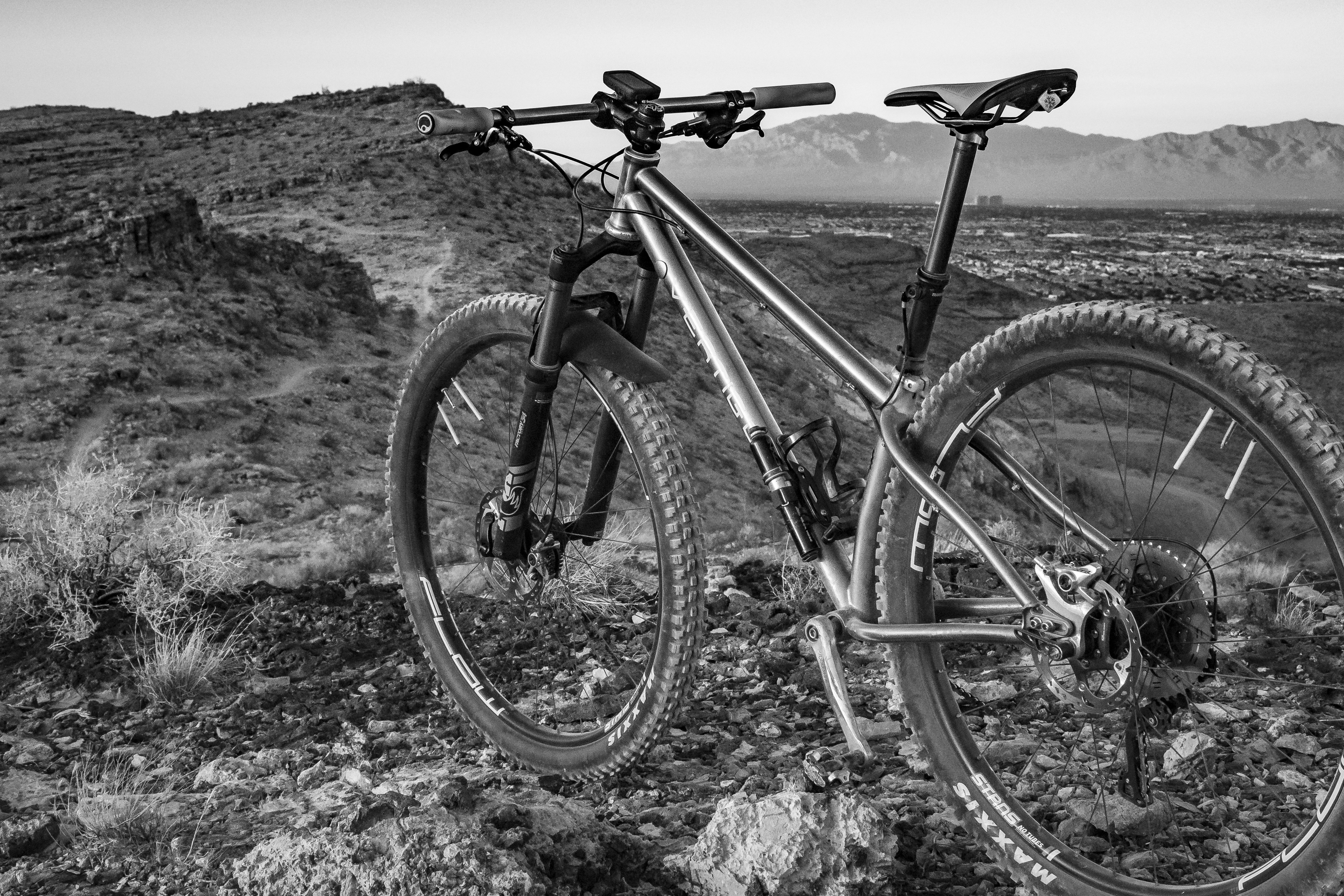 A sleek mountain bike positioned on rocky terrain, showcasing its design against a vast, arid landscape. The monochrome tones emphasize the ruggedness of the environment.