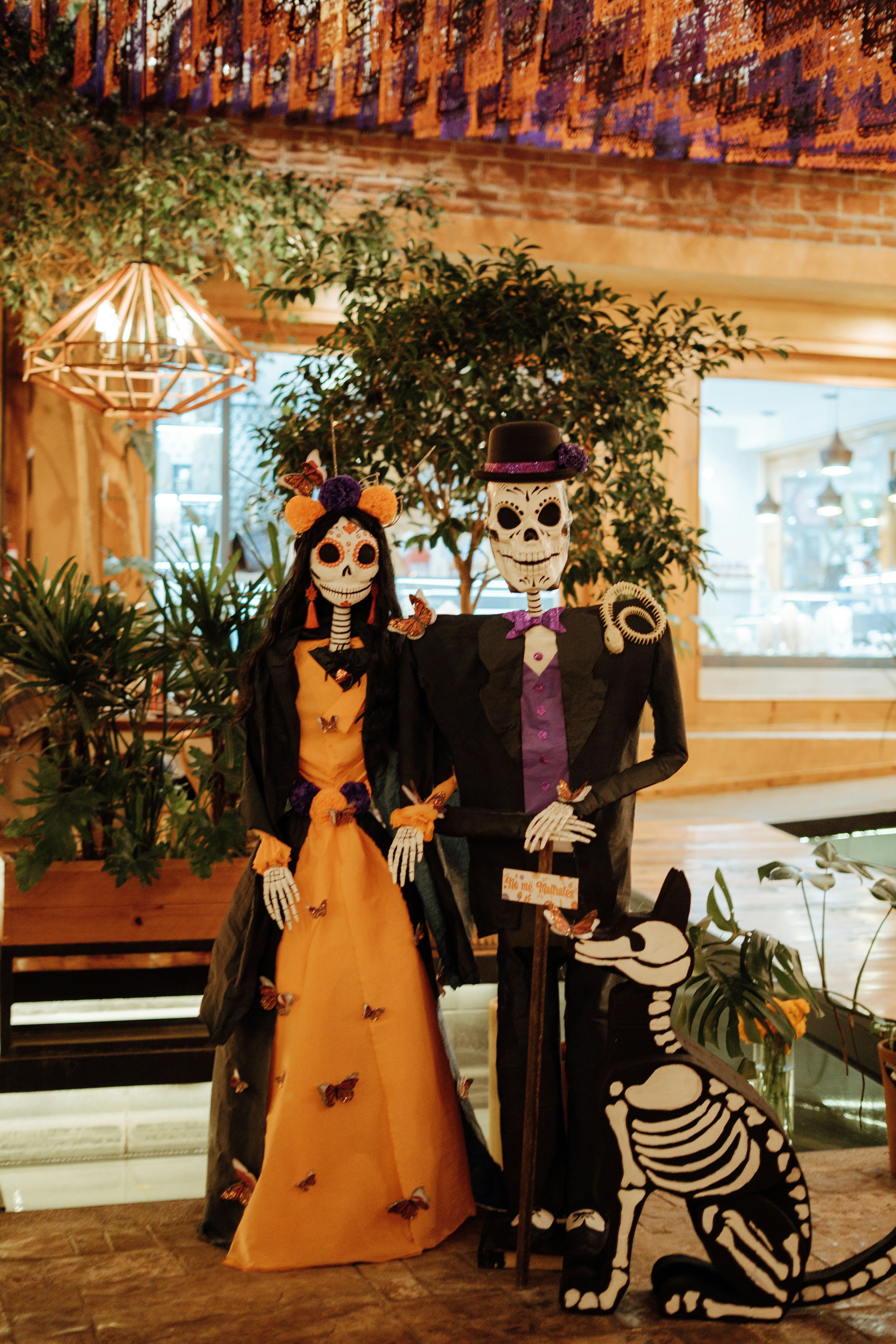 Colorful skeleton figures dressed in traditional attire, surrounded by lush greenery and festive decorations. A playful dog skeleton adds to the scene.