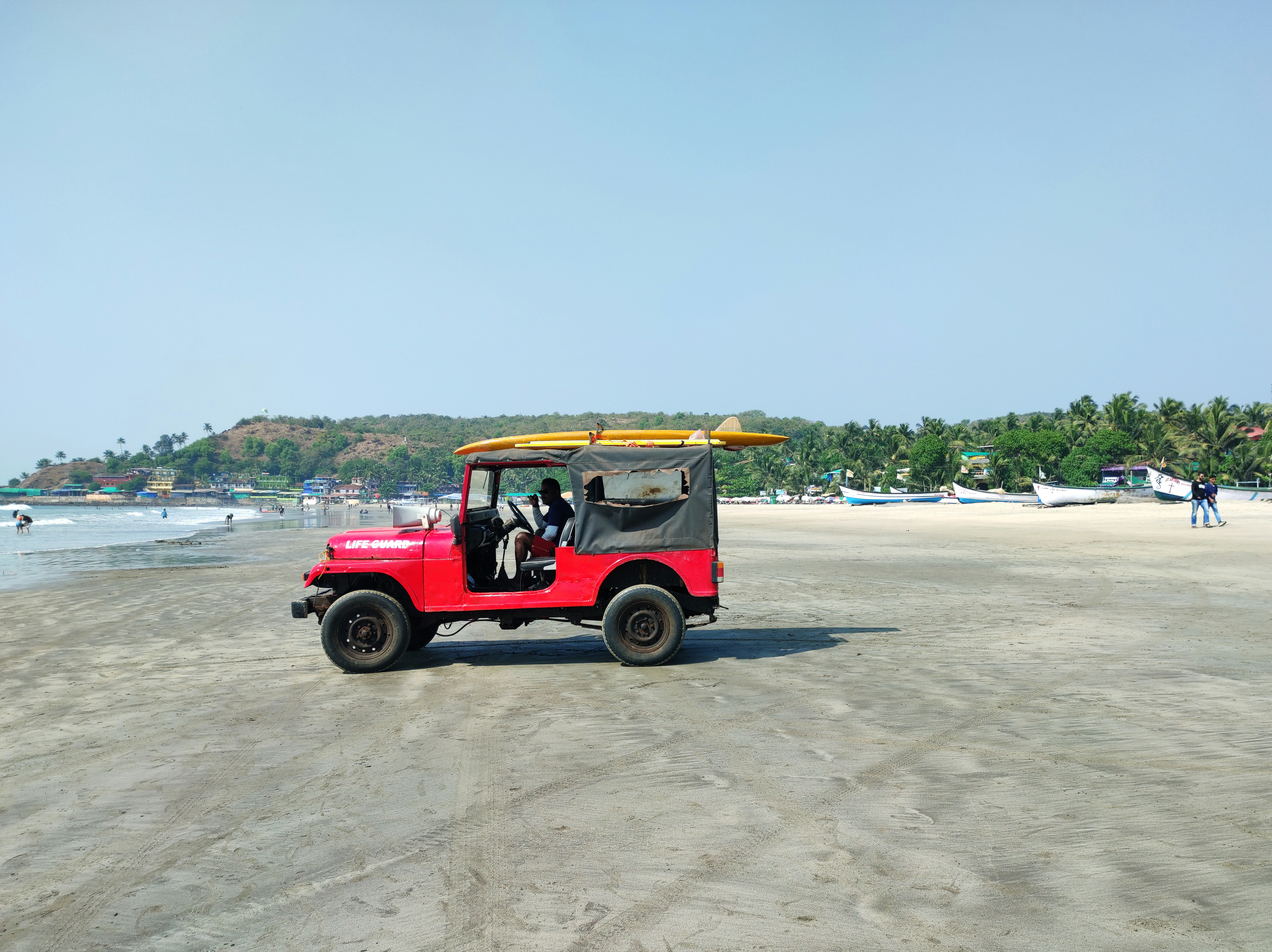 Red jeep parked on a sandy beach with a backdrop of palm trees and fishing boats. The scene captures a moment of relaxation by the coast.