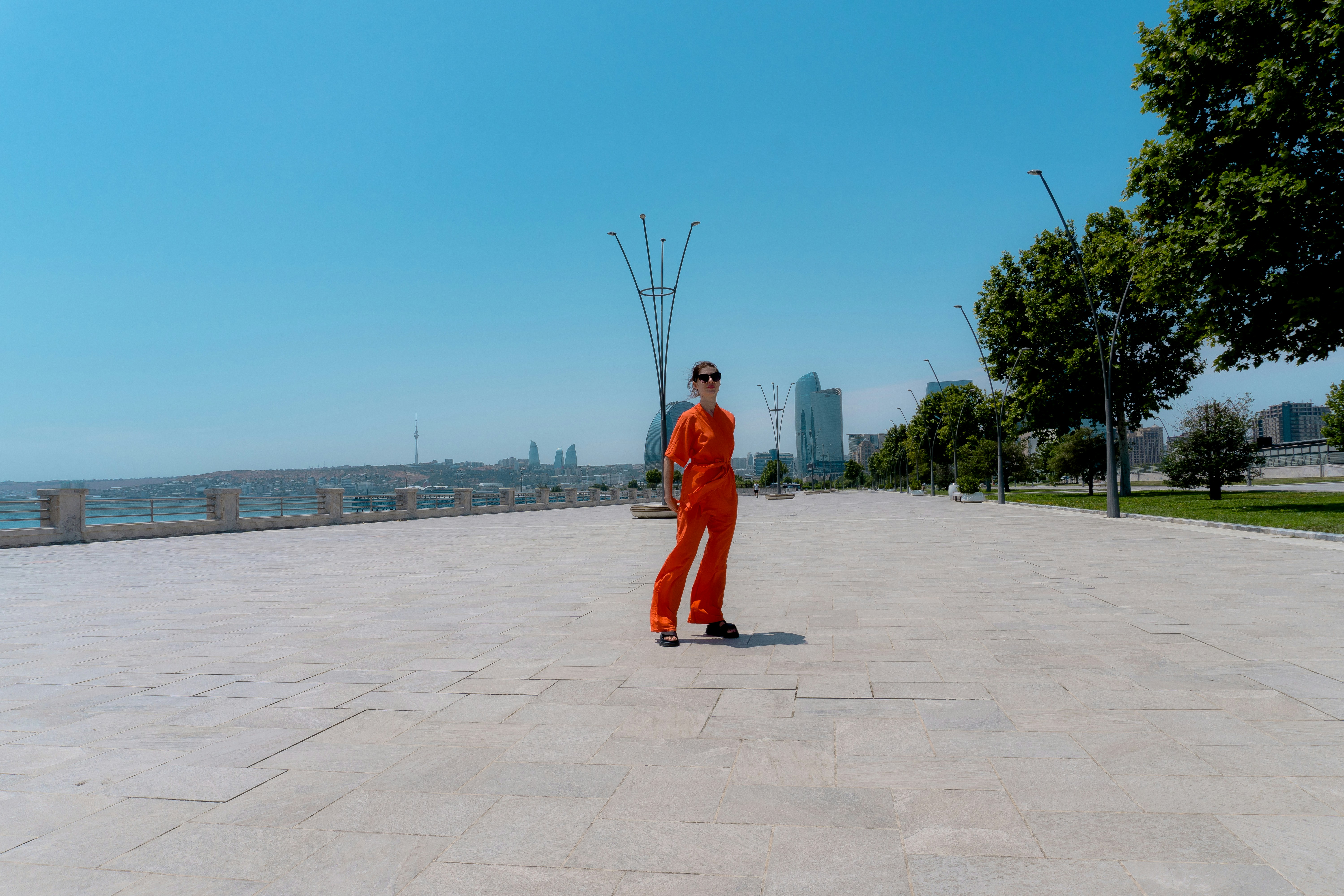 Crimson-clad figure anchors a sunlit waterfront promenade with a distant skyline