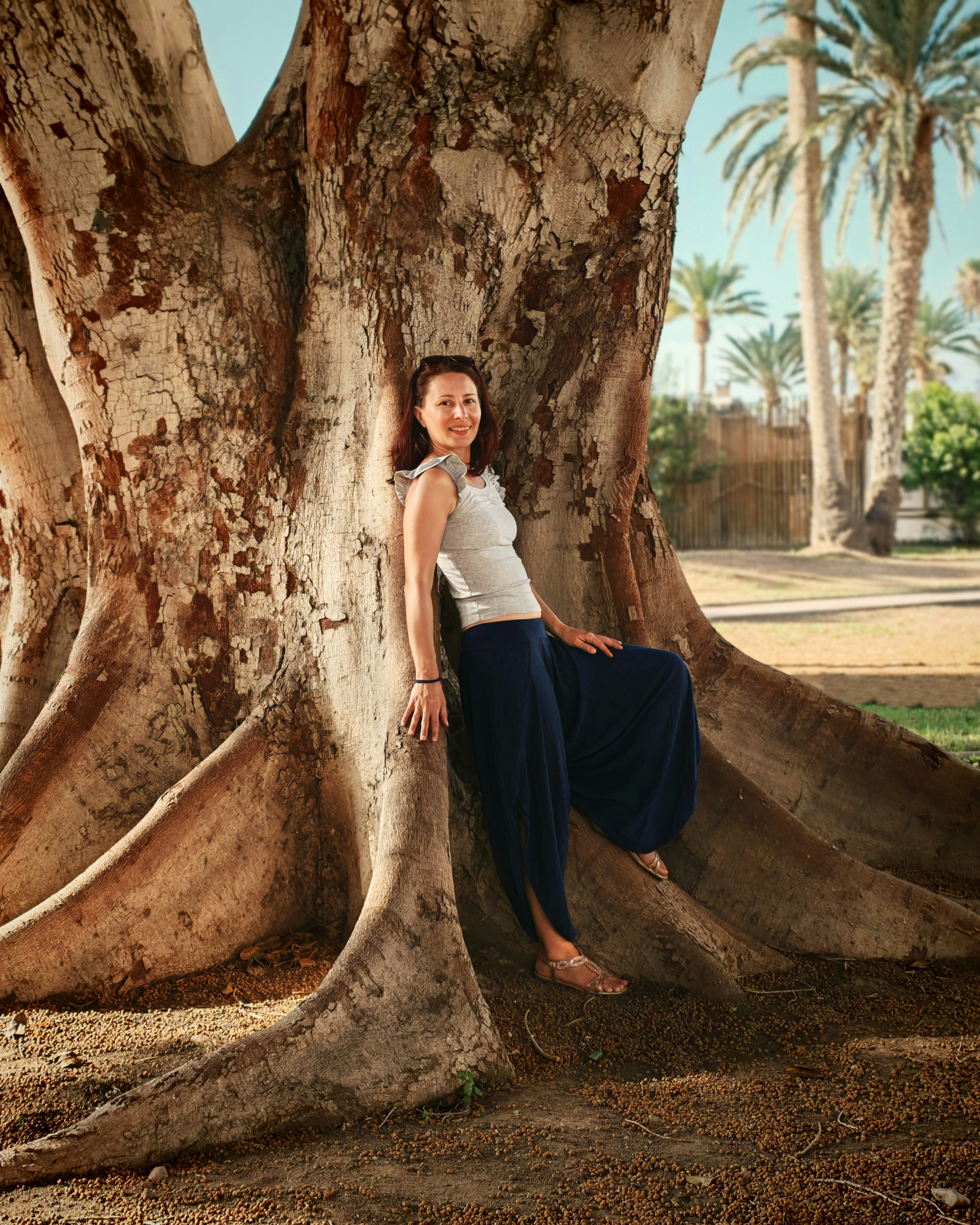 Woman leaning against a large tree trunk, surrounded by palm trees in a sunlit park. The scene conveys a sense of tranquility and connection to nature.