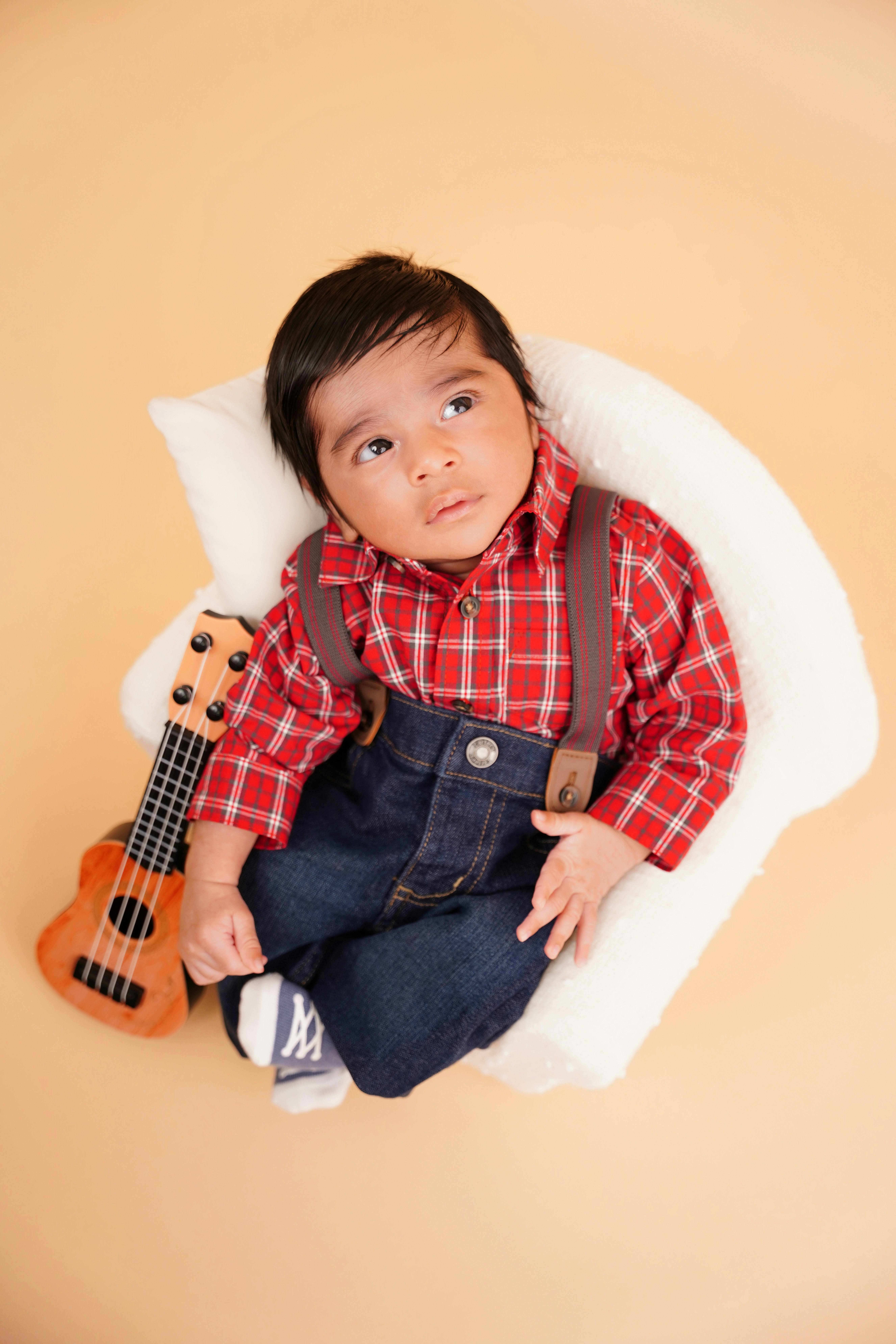 A young child in a plaid shirt and denim overalls sits in a cozy chair, gazing thoughtfully while holding a ukulele. The warm background complements the playful scene.