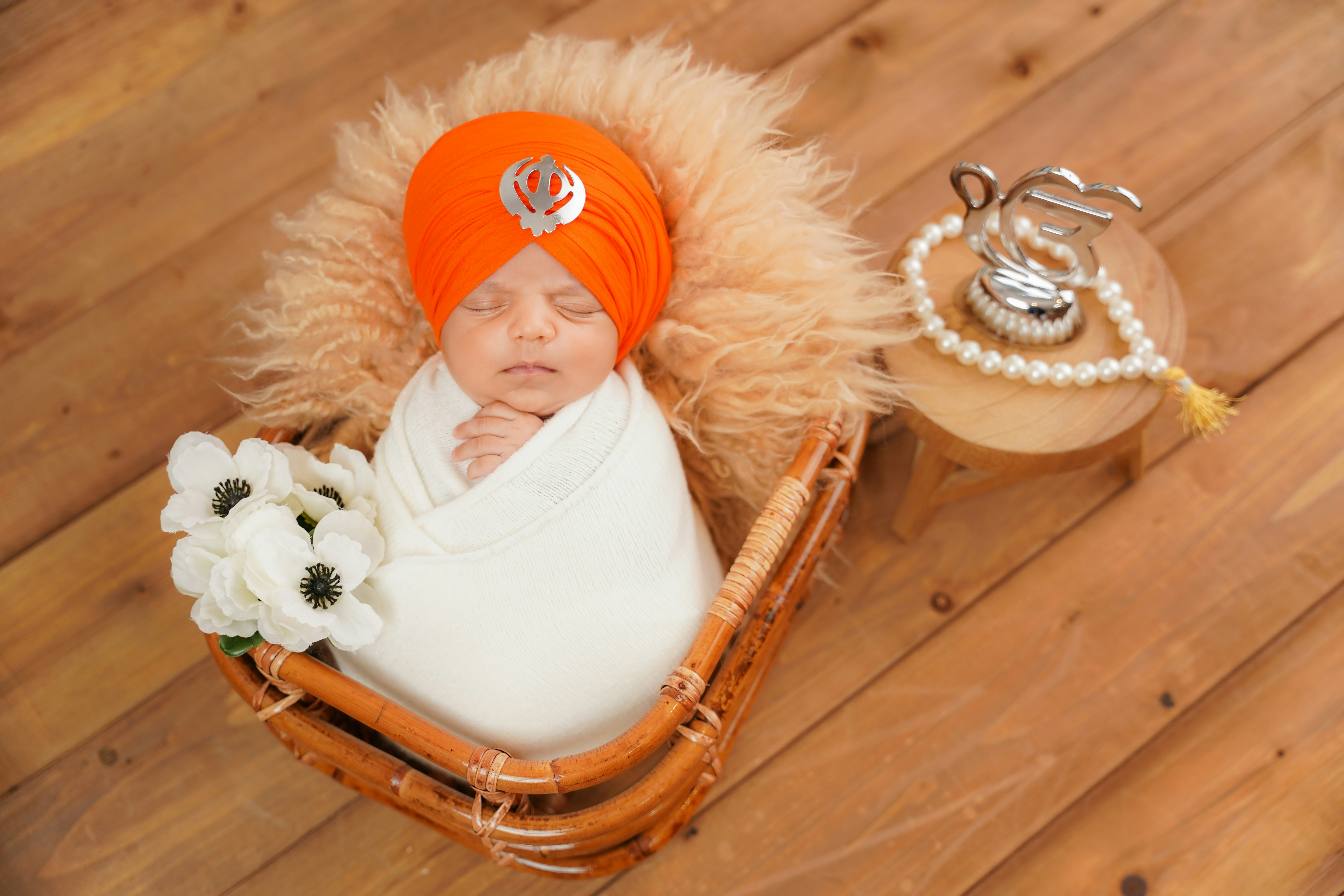 Infant swaddled in a white blanket, adorned with an orange turban, nestled in a woven basket surrounded by flowers and decorative items. A symbol of cultural heritage is present.