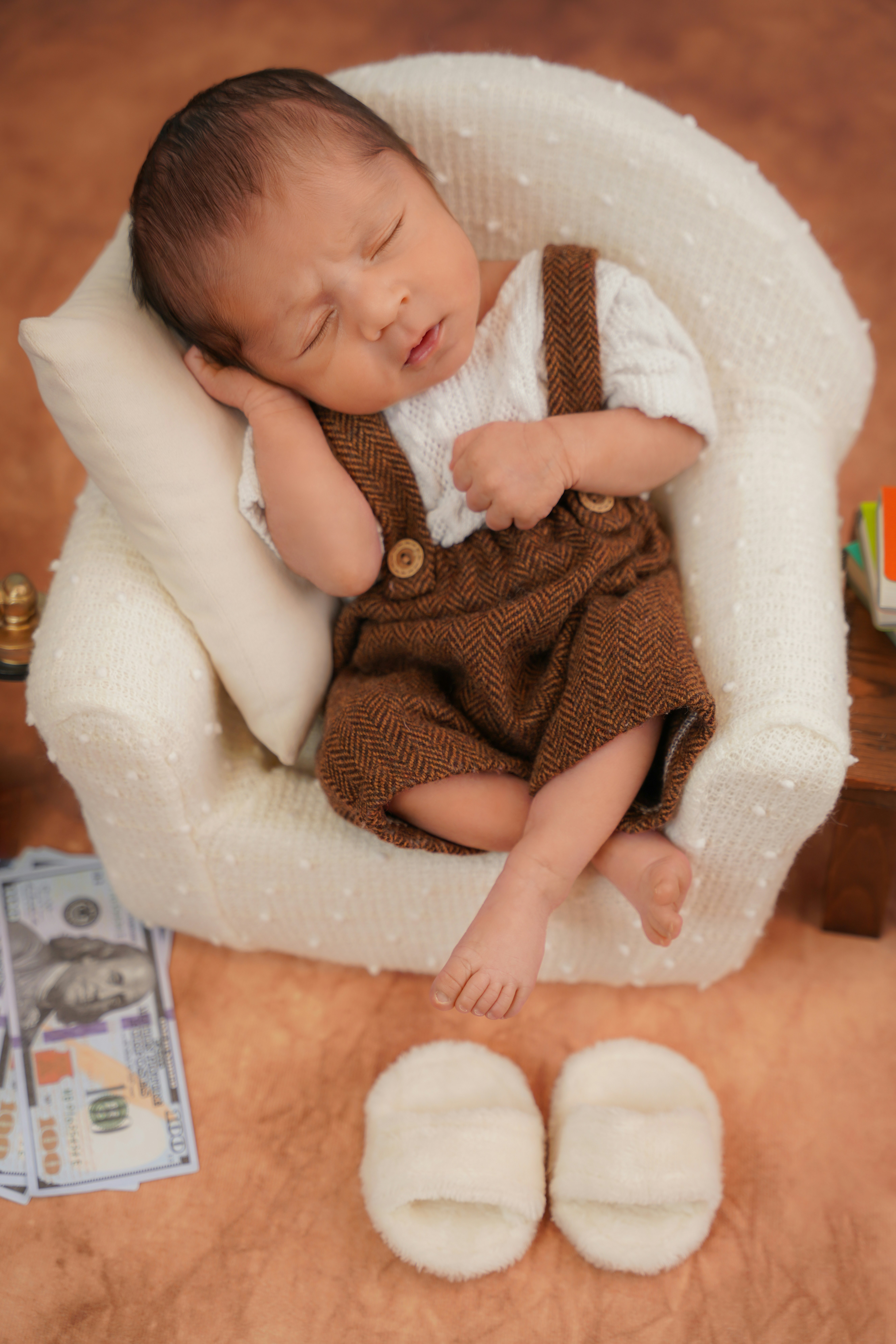 A peacefully sleeping baby nestled in a plush chair, adorned in brown overalls, with soft slippers nearby and dollar bills scattered on the floor.