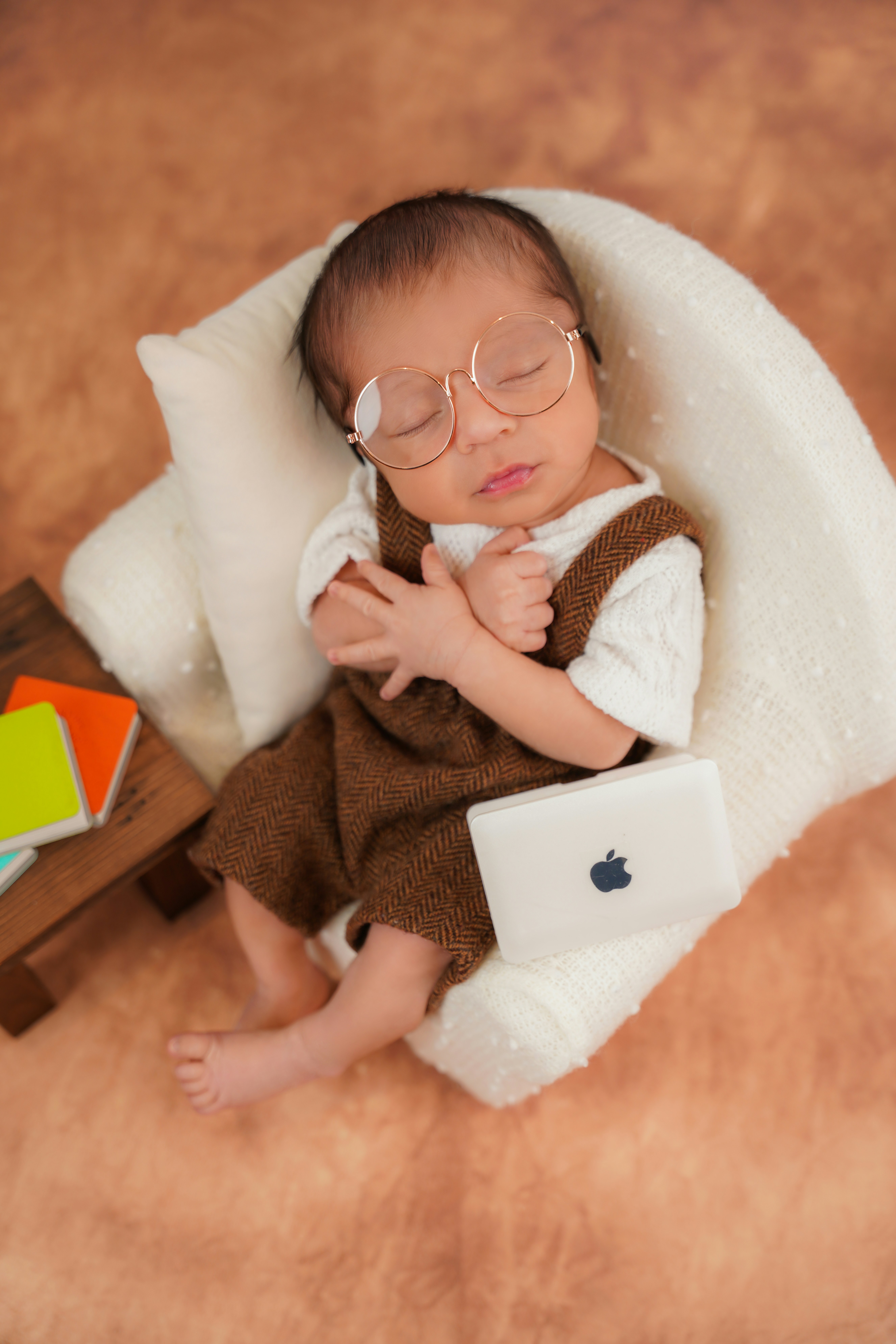 Adorable baby dressed in a brown outfit and oversized glasses, peacefully napping in a cozy chair with a small laptop and colorful notebooks nearby.