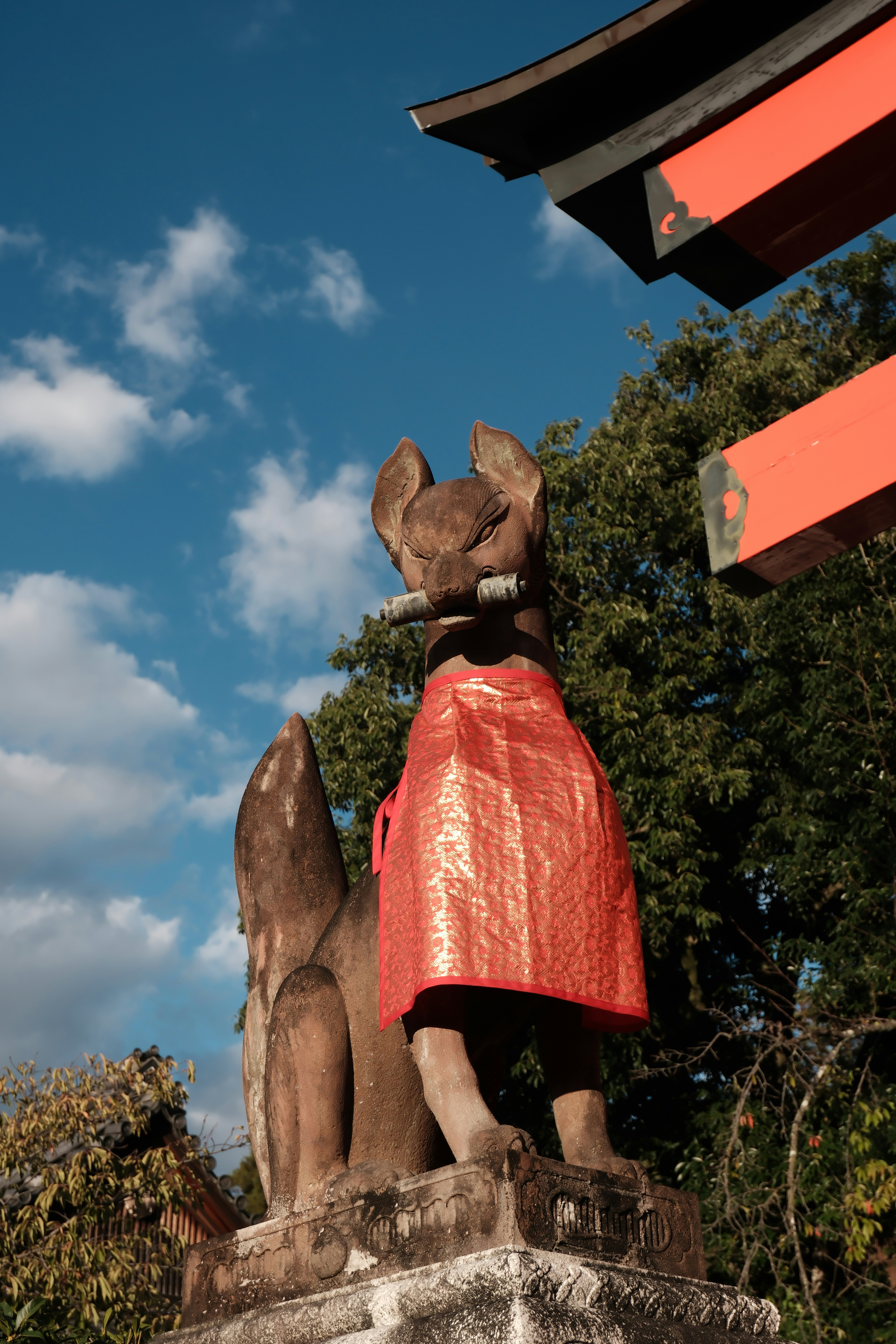 A stone fox statue adorned with a vibrant red cloth, standing watch beneath a bright blue sky and scattered clouds.