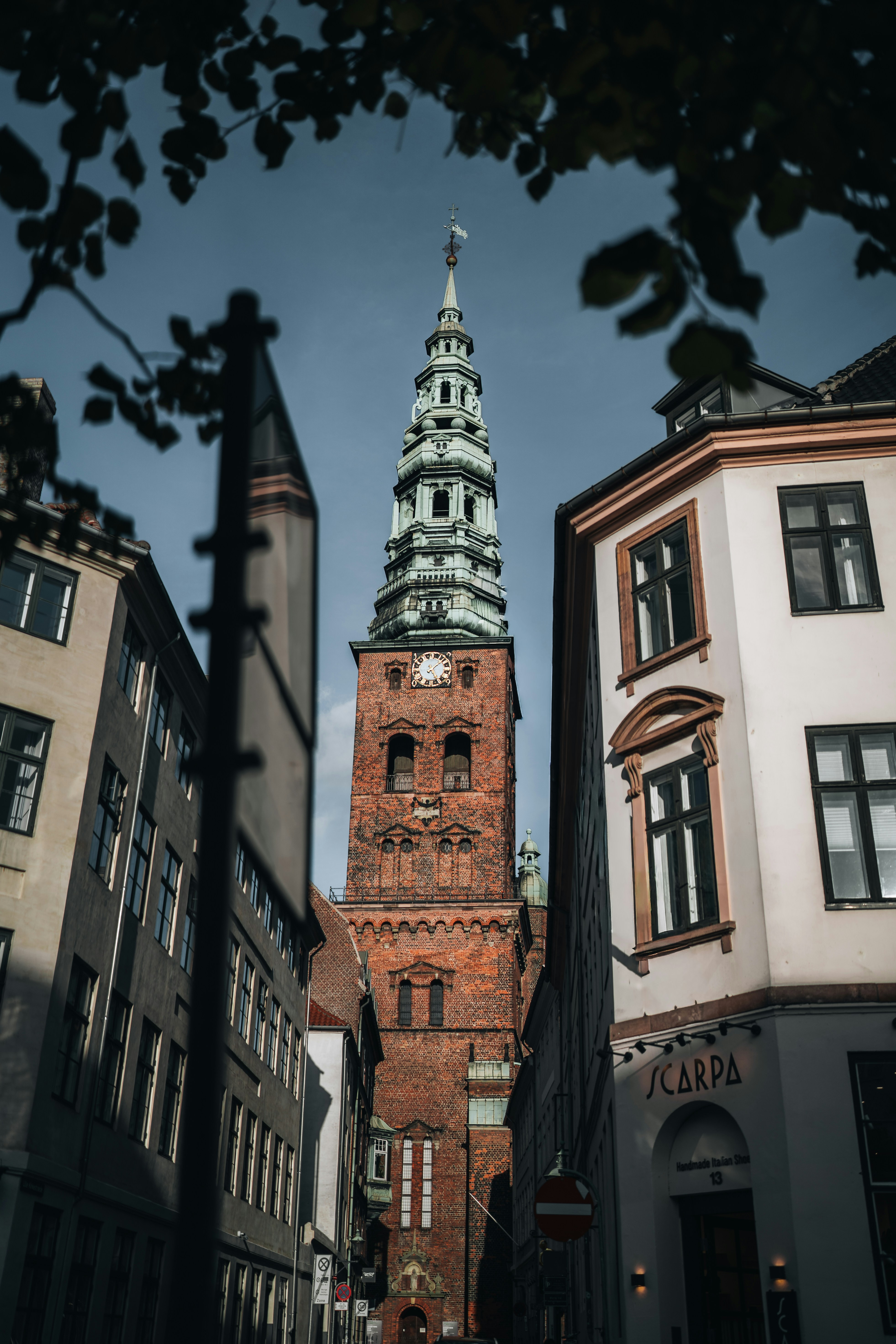 Tall brick church tower framed by buildings and leaves