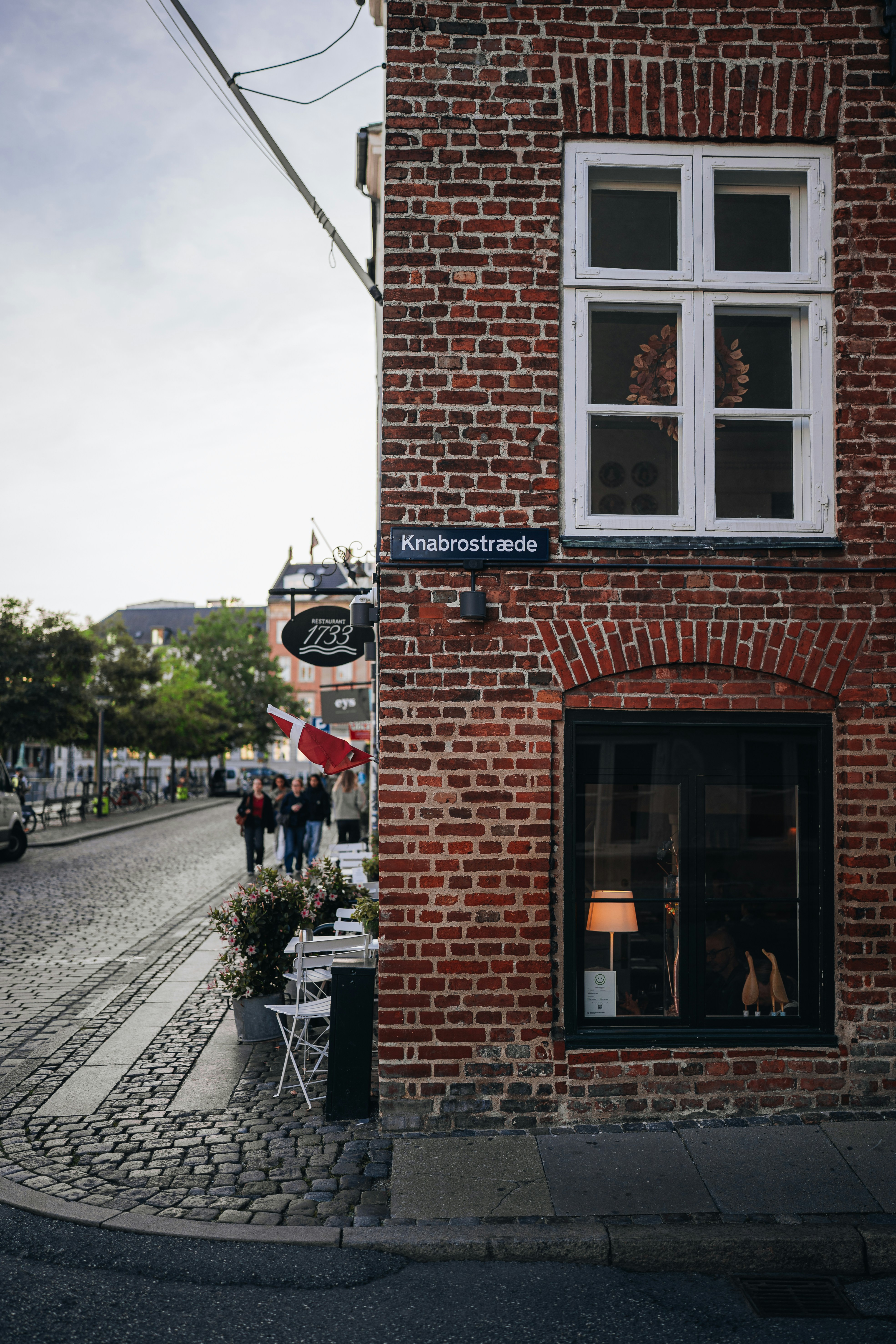 Brick facade of a cozy corner building with a lamp visible through the window, located on Knabrostrae street. A glimpse of pedestrians and outdoor seating adds to the urban atmosphere.