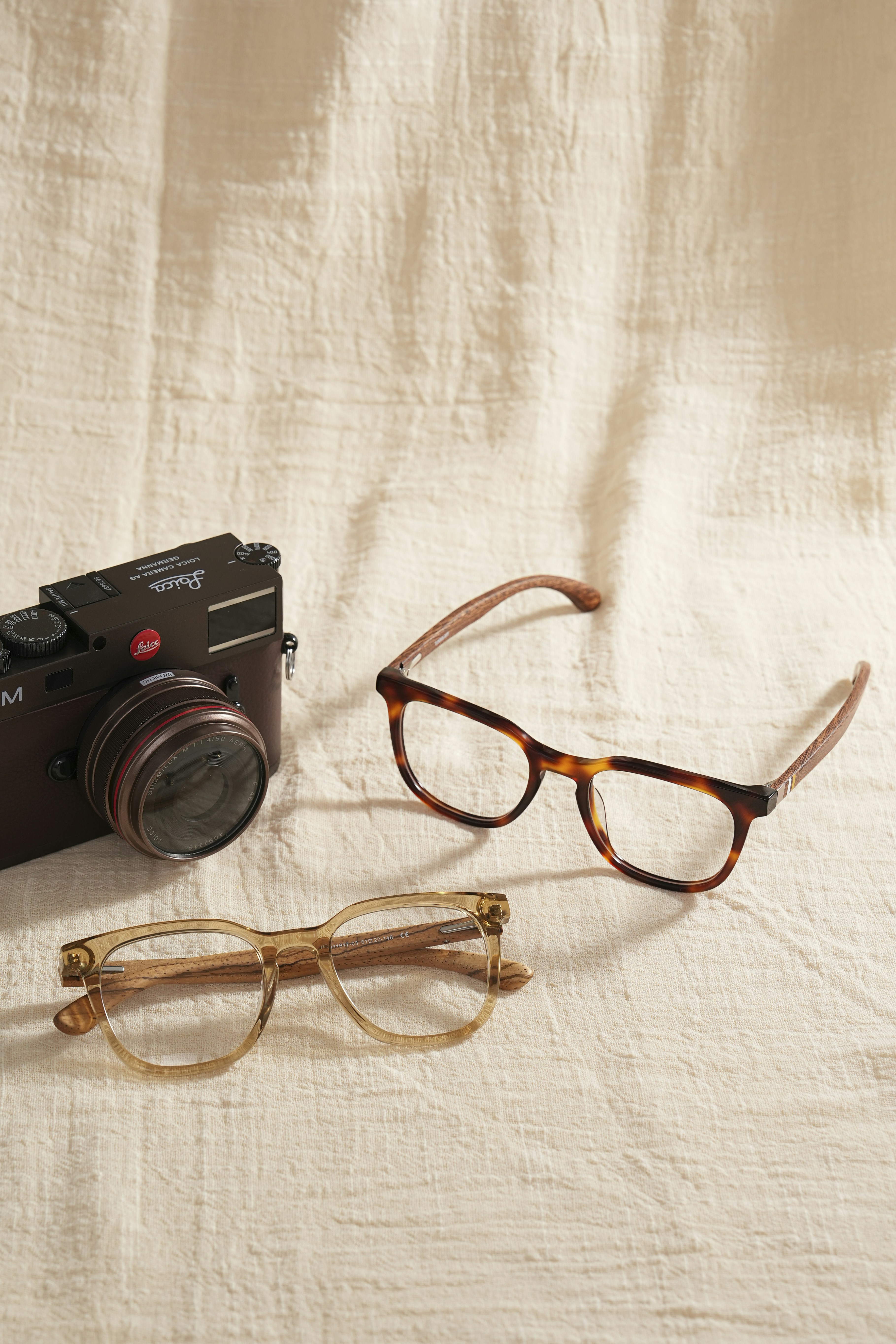 Two Vooglam square eyeglasses—tortoiseshell and champagne clear—styled beside a camera on natural linen for a cozy lifestyle mood.