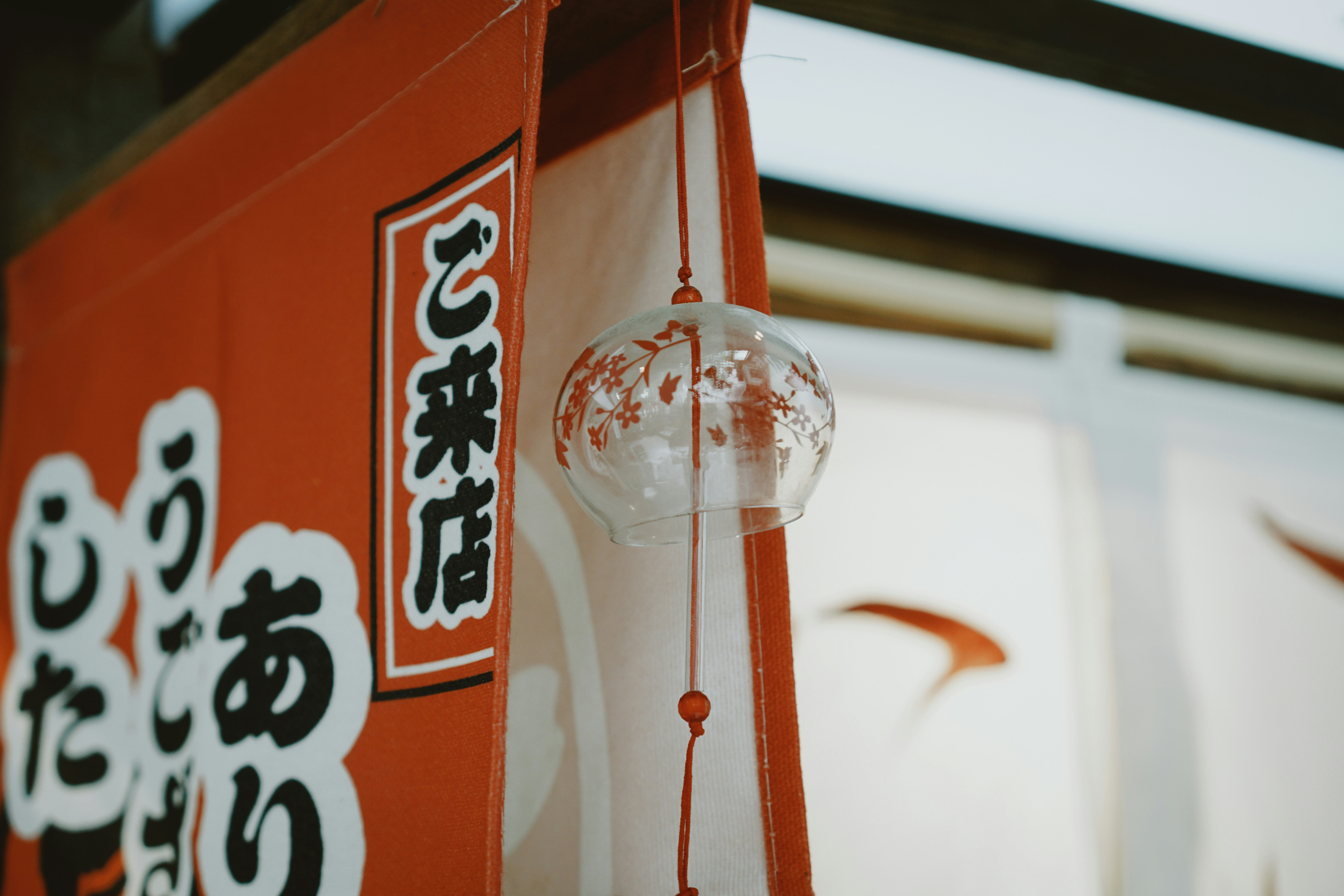 Delicate glass wind chime hanging next to vibrant red fabric banners adorned with Japanese characters. The scene evokes a sense of cultural heritage.