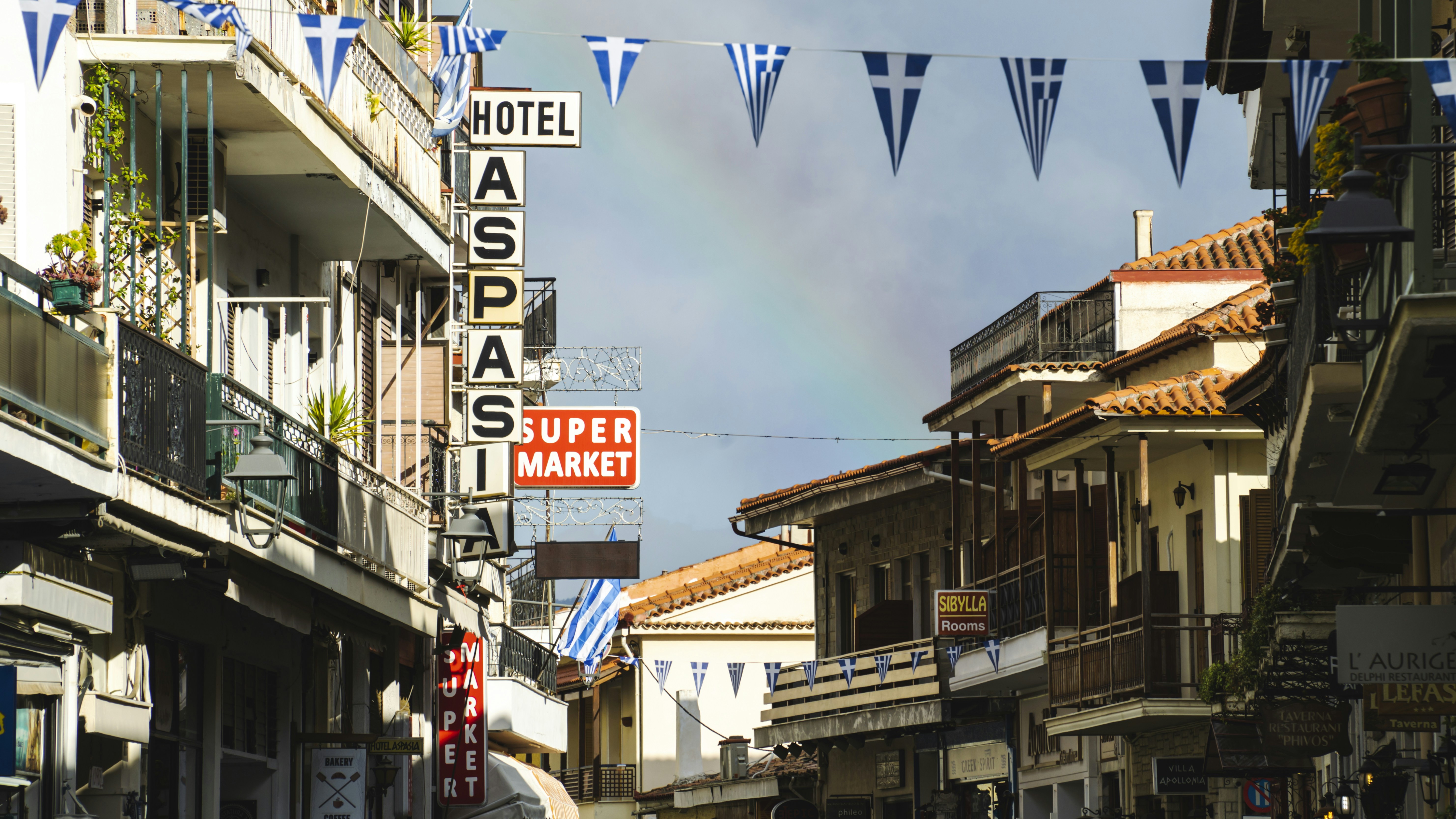 A vibrant street scene in Aspas, featuring local shops and festive decorations, with Greek flags adorning the buildings. The atmosphere reflects a lively community spirit.