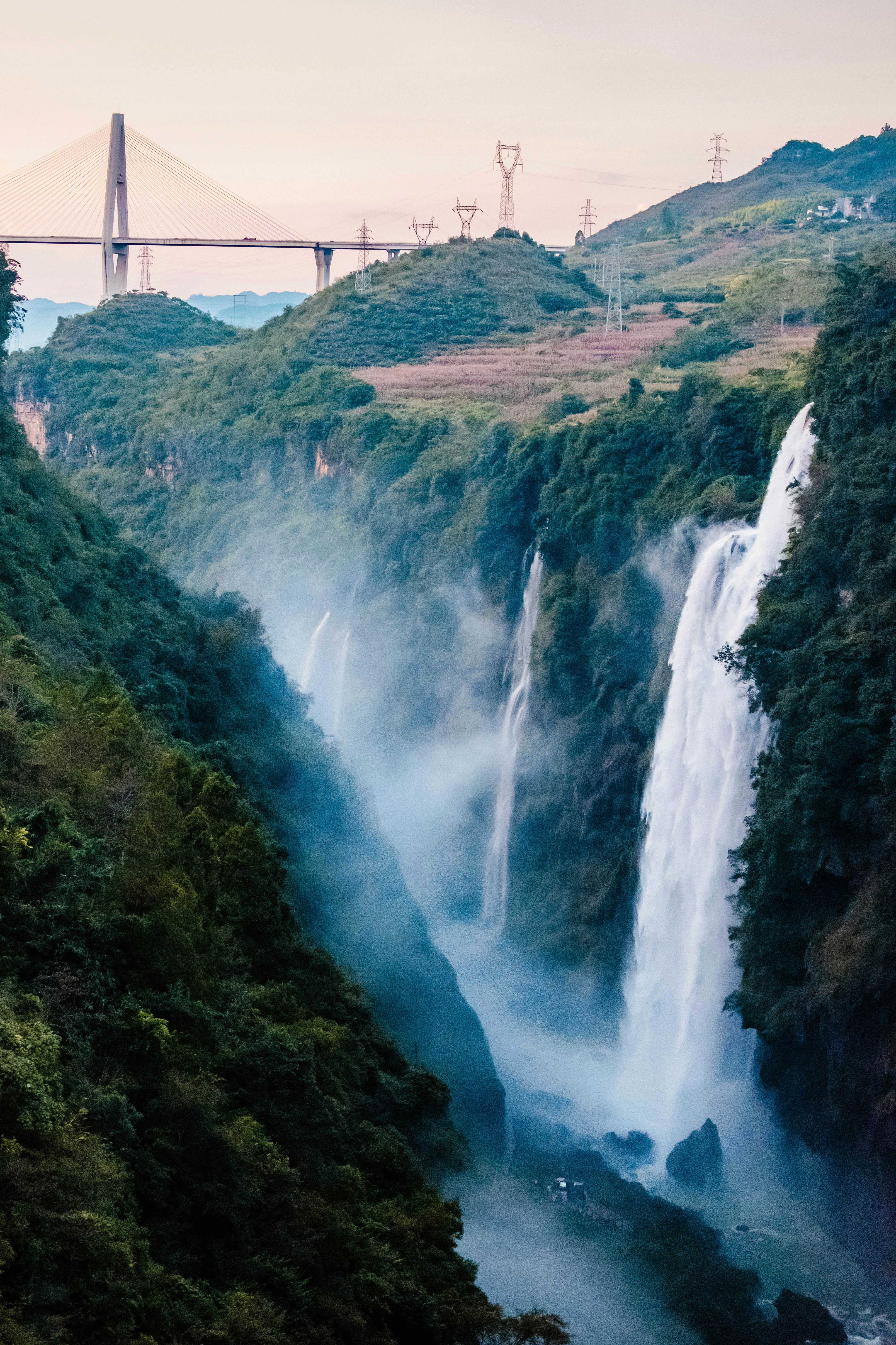 Majestic waterfalls cascading into a misty gorge, framed by a modern bridge and lush greenery. The scene captures the harmony between nature and engineering.