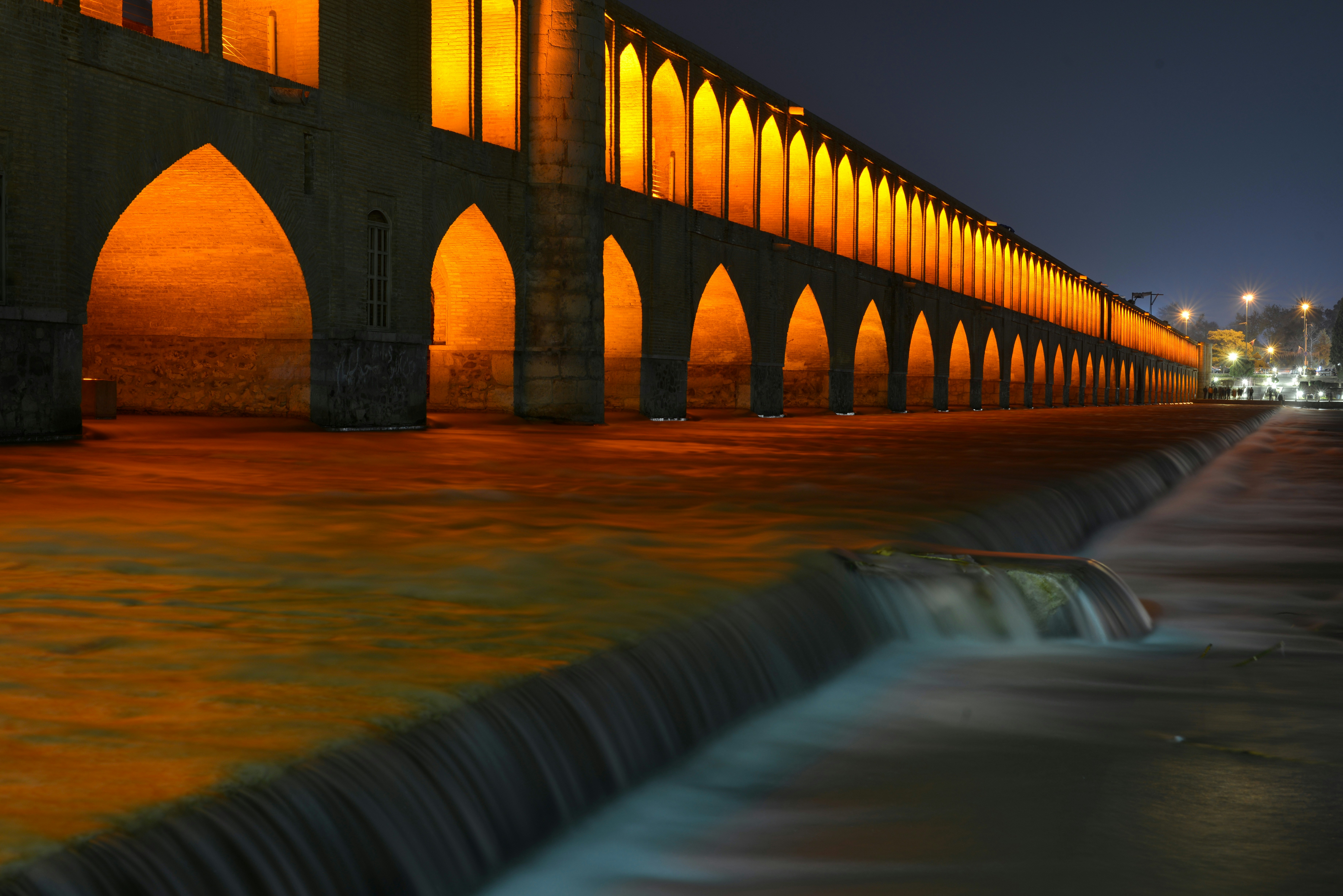 A long-exposure night shot of Si-o-se-pol (33 Bridge) in Isfahan, Iran — a stunning example of Persian architecture and engineering. The arches reflected in the Zayandeh River create a mesmerizing symmetry, blending centuries-old craftsmanship with the serene beauty of night. Captured in stillness, this image celebrates cultural heritage, light, and the timeless dialogue between water and stone. | A long bridge illuminated at night over water