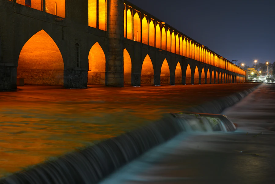 Historic stone bridge with illuminated arches casting orange reflections across a flowing river at night.