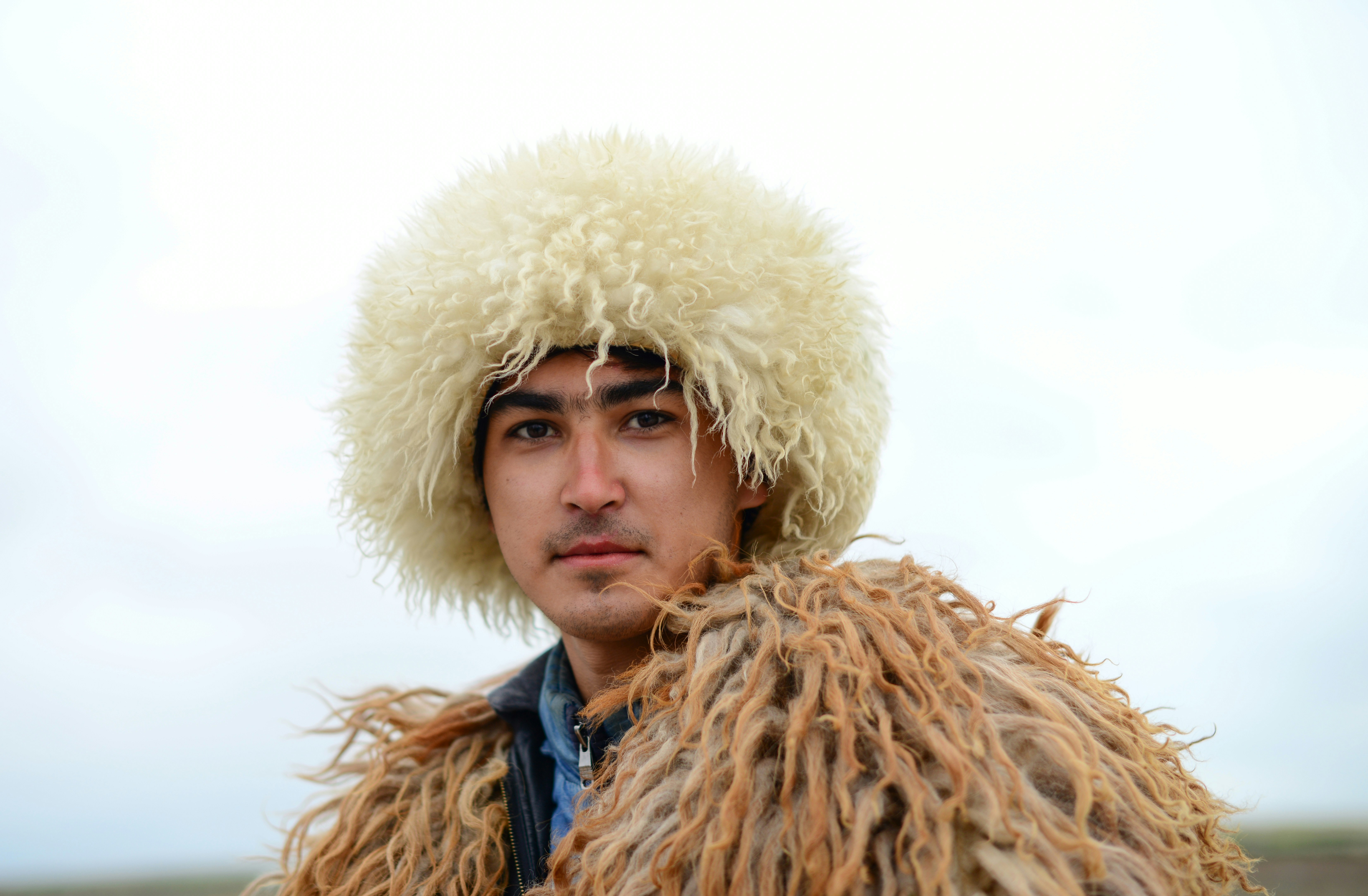 A portrait of a Turkmen man, wearing a traditional cream-colored wool hat and brown sheepskin coat, capturing the rich cultural heritage of Turkmen people. The texture of the wool and the warmth of the clothing highlight centuries-old traditions, while the subject’s gaze conveys a sense of quiet strength and timeless character. Captured in natural light to preserve authenticity, this portrait celebrates human stories, tradition, and cultural identity. | Man wearing a fluffy hat and fur coat.