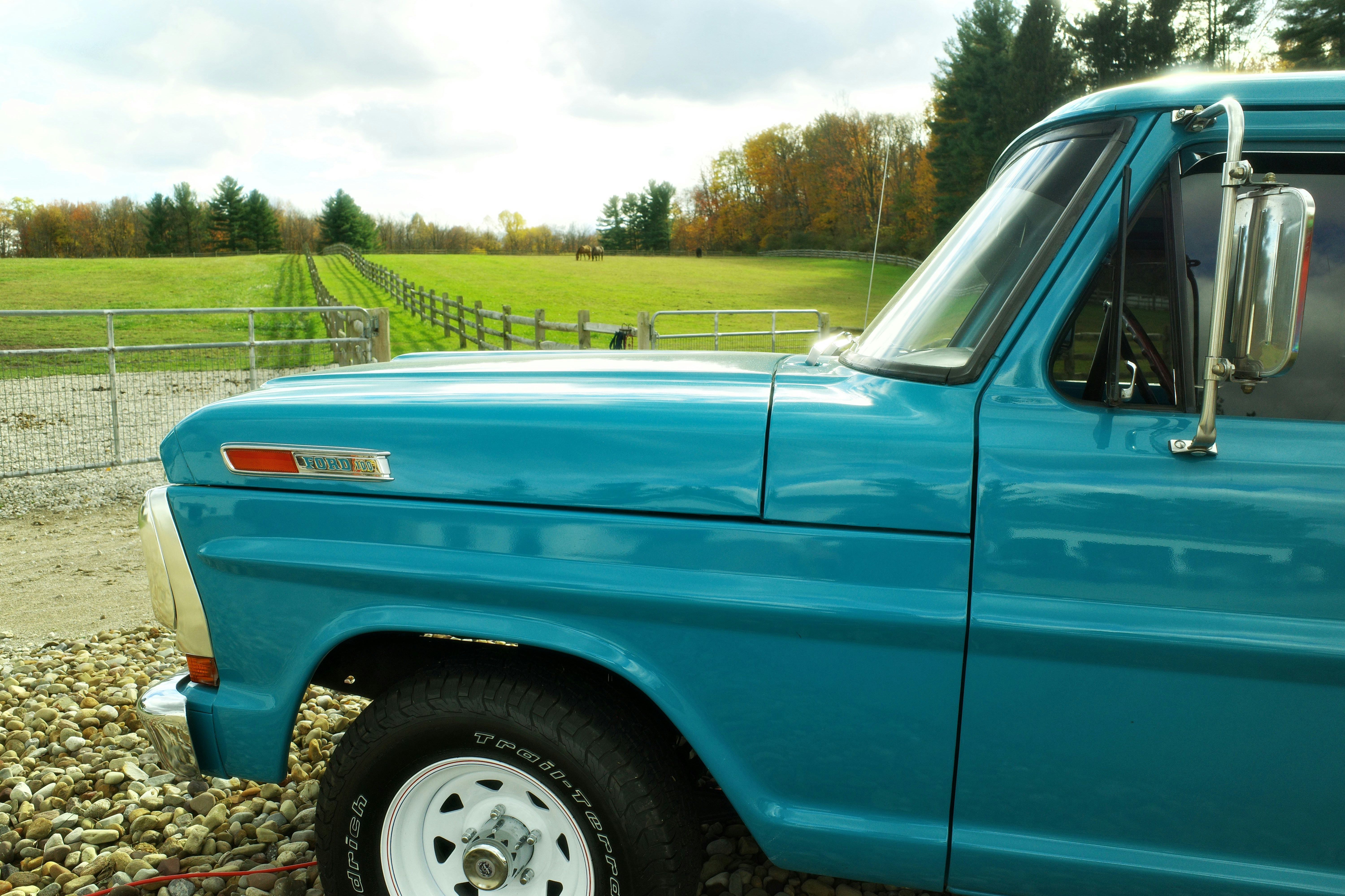 Classic turquoise pickup truck parked beside a picturesque field with a fence and autumn foliage in the background.