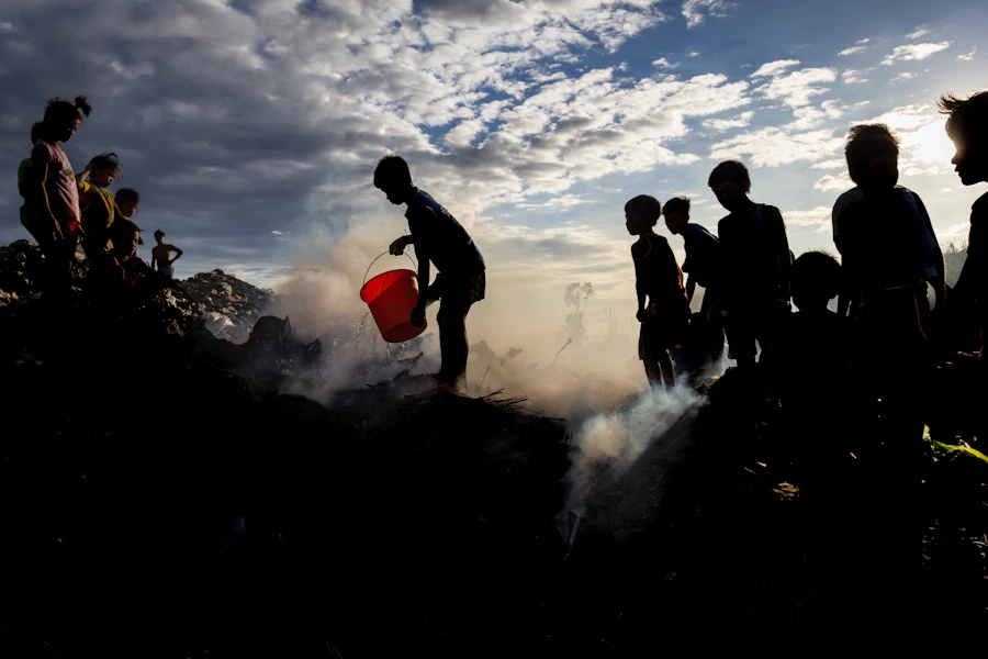 Backlit group of children or workers stands on a smoky hillside, with one person holding a bright red bucket. Dramatic clouds and low sun turn the figures into strong silhouettes.