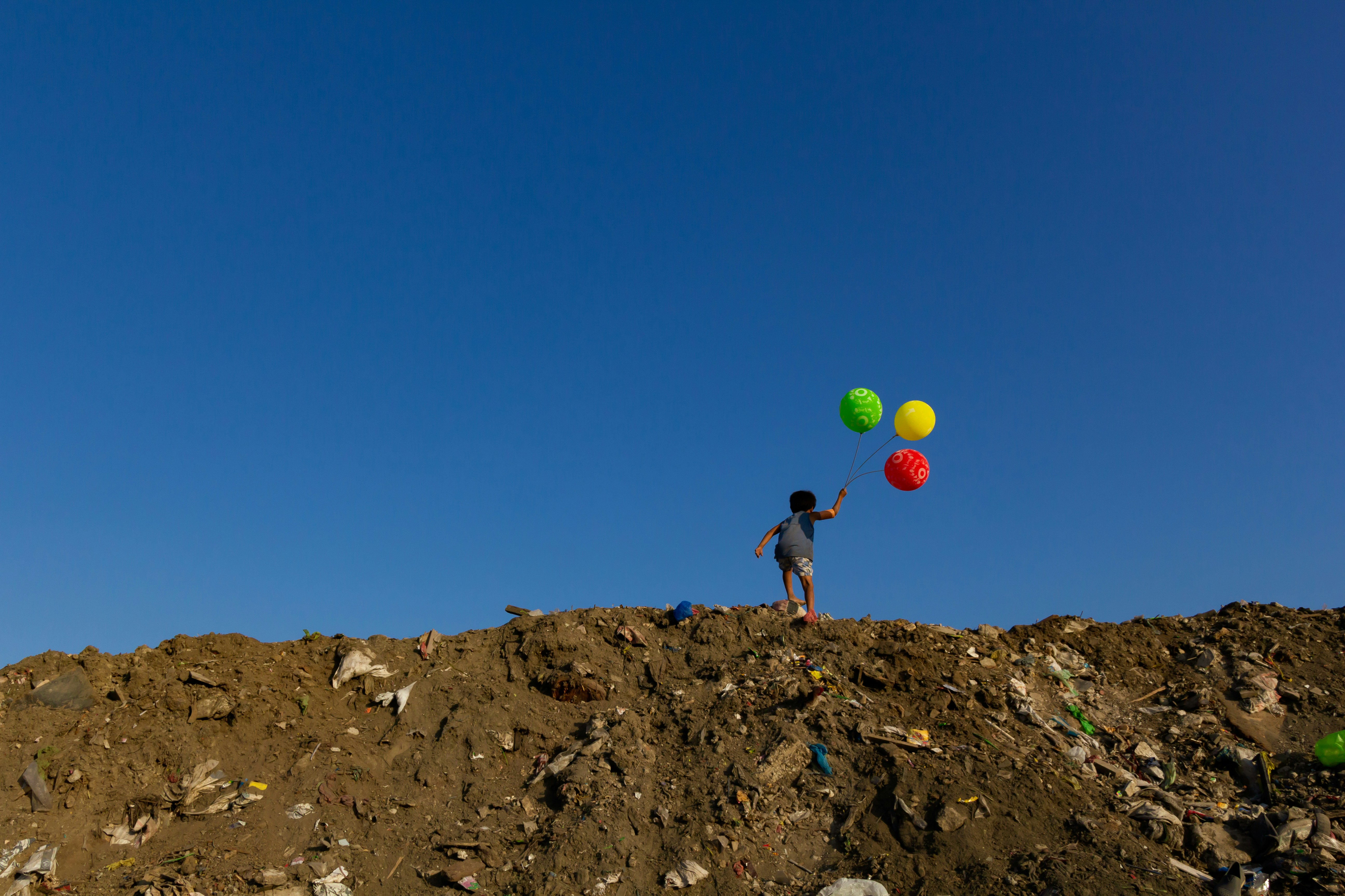 Child standing on a mound of debris, holding colorful balloons against a clear blue sky.