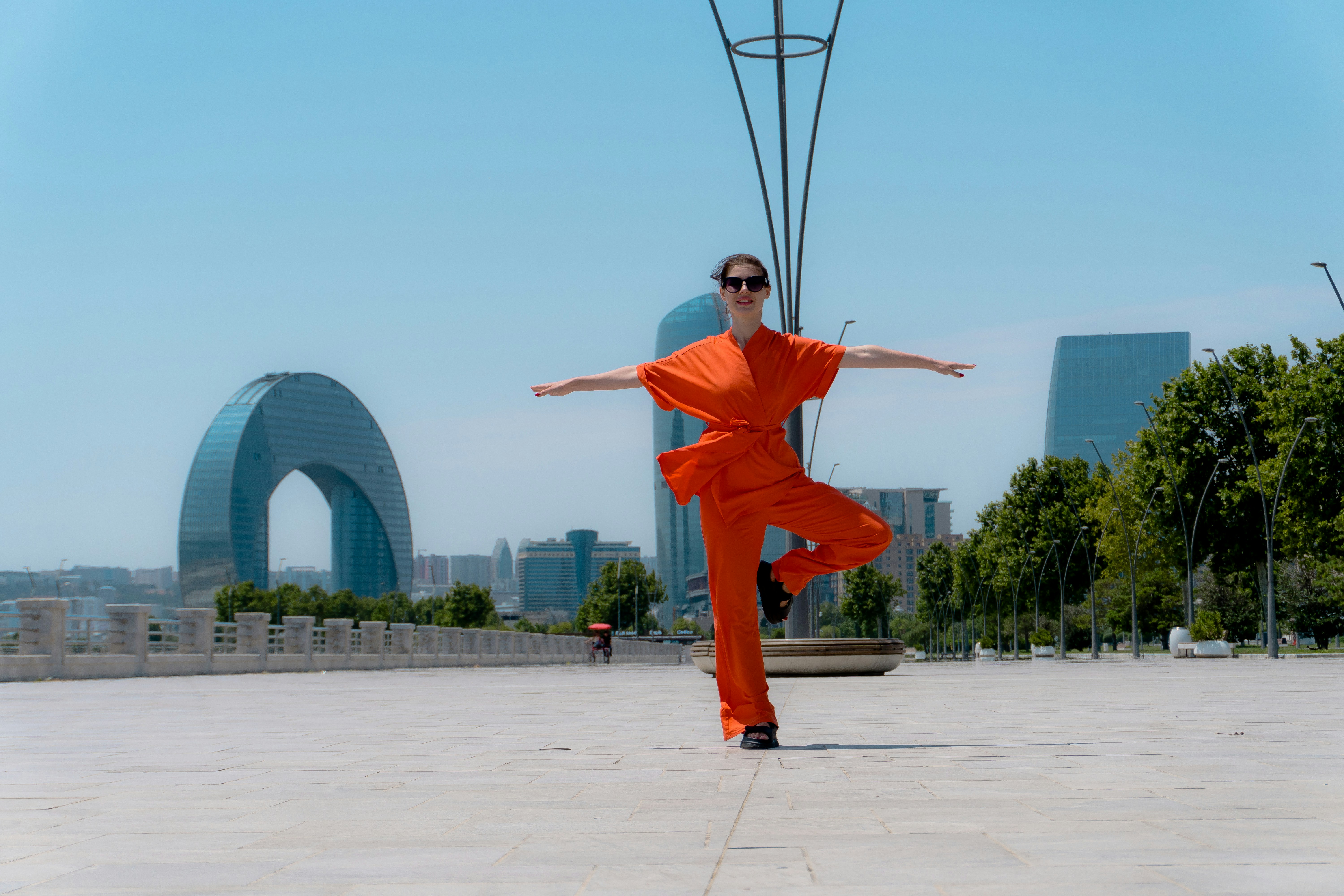 Orange-clad dancer balances mid-air on a sunlit plaza with modern skyline in the background