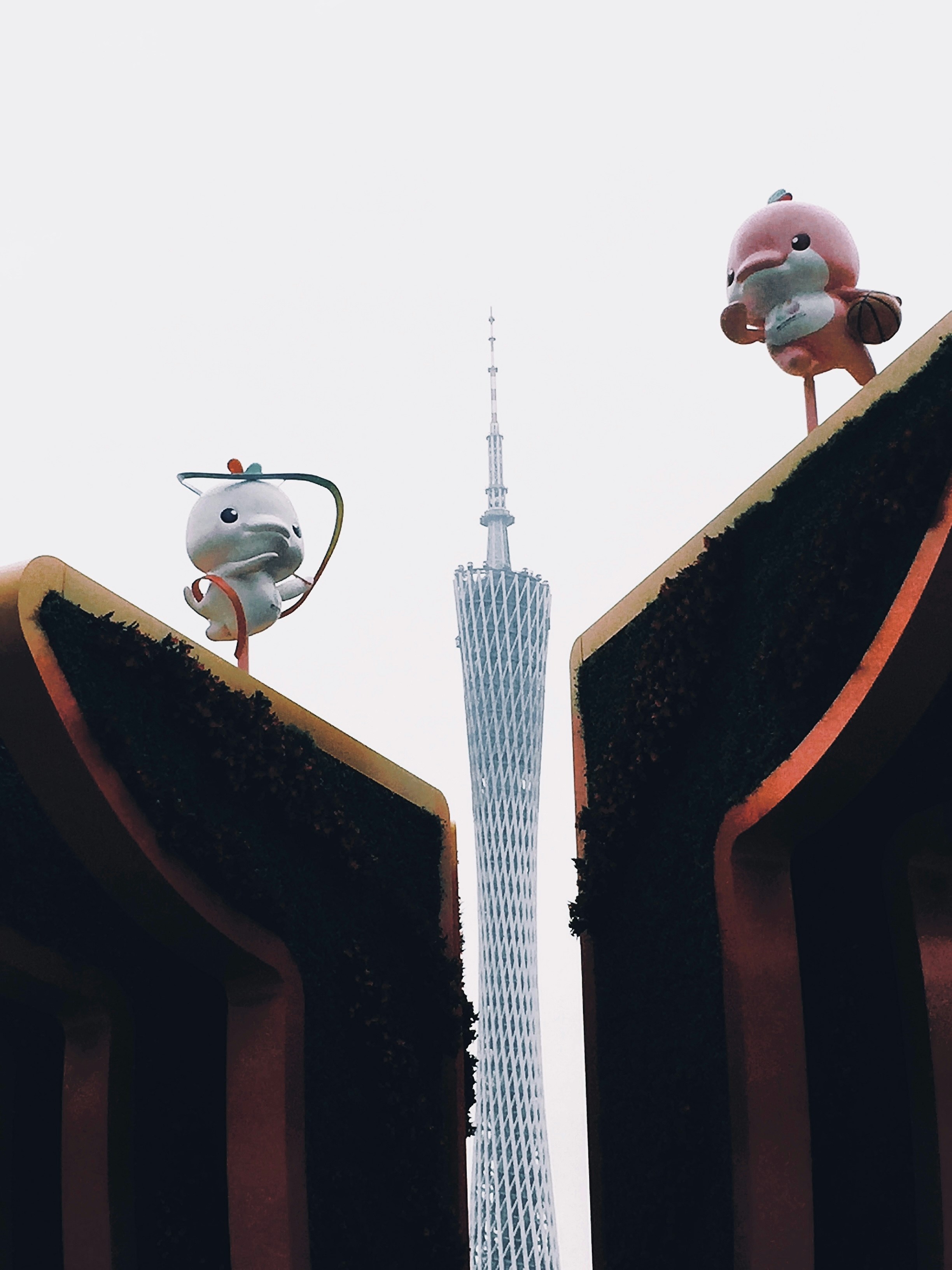 Two playful cartoon characters perched atop vibrant structures, framing the towering skyline of a modern city landmark.