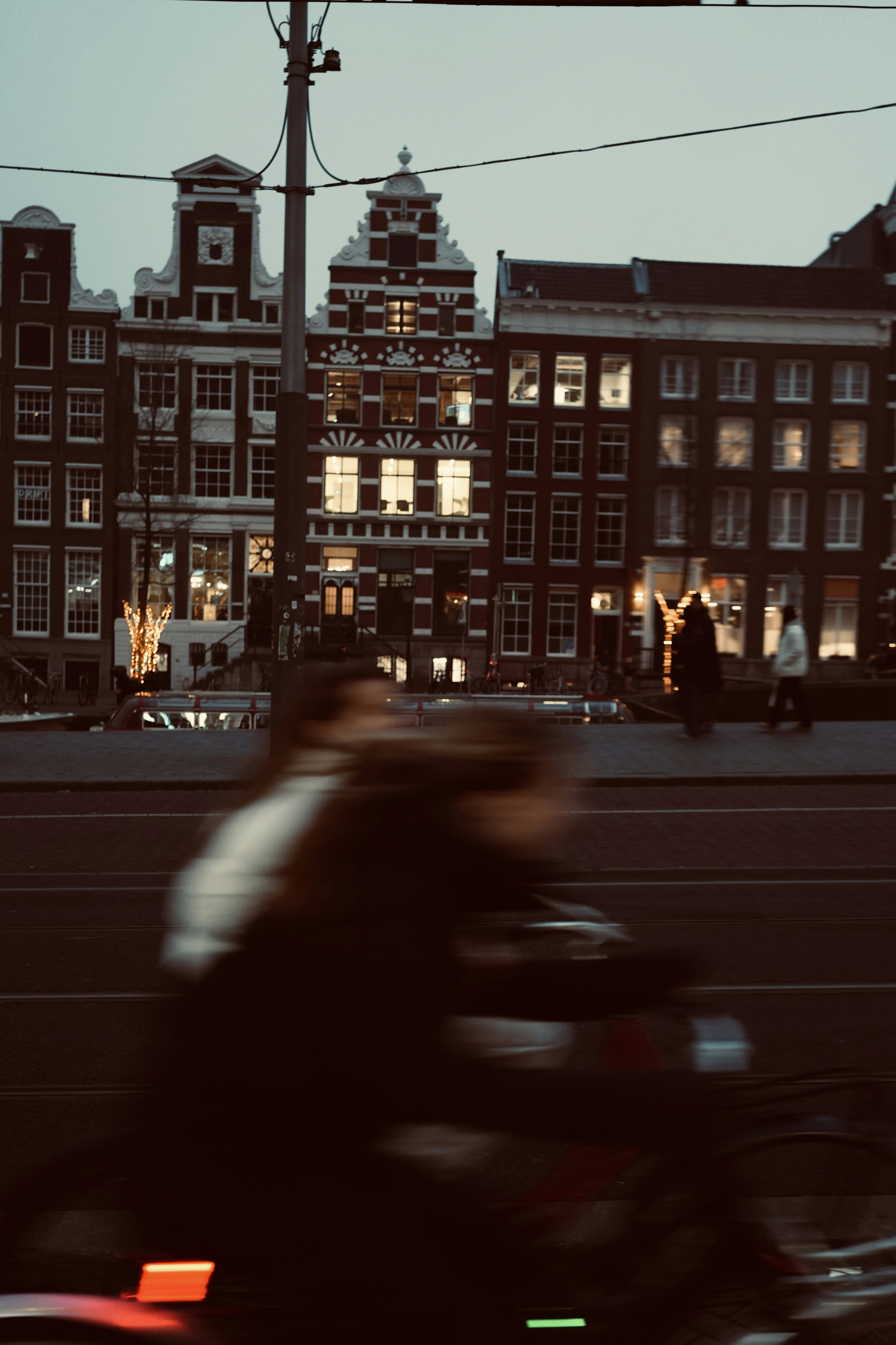 Two cyclists blur past historic buildings adorned with soft lights, capturing the dynamic essence of city life at dusk.