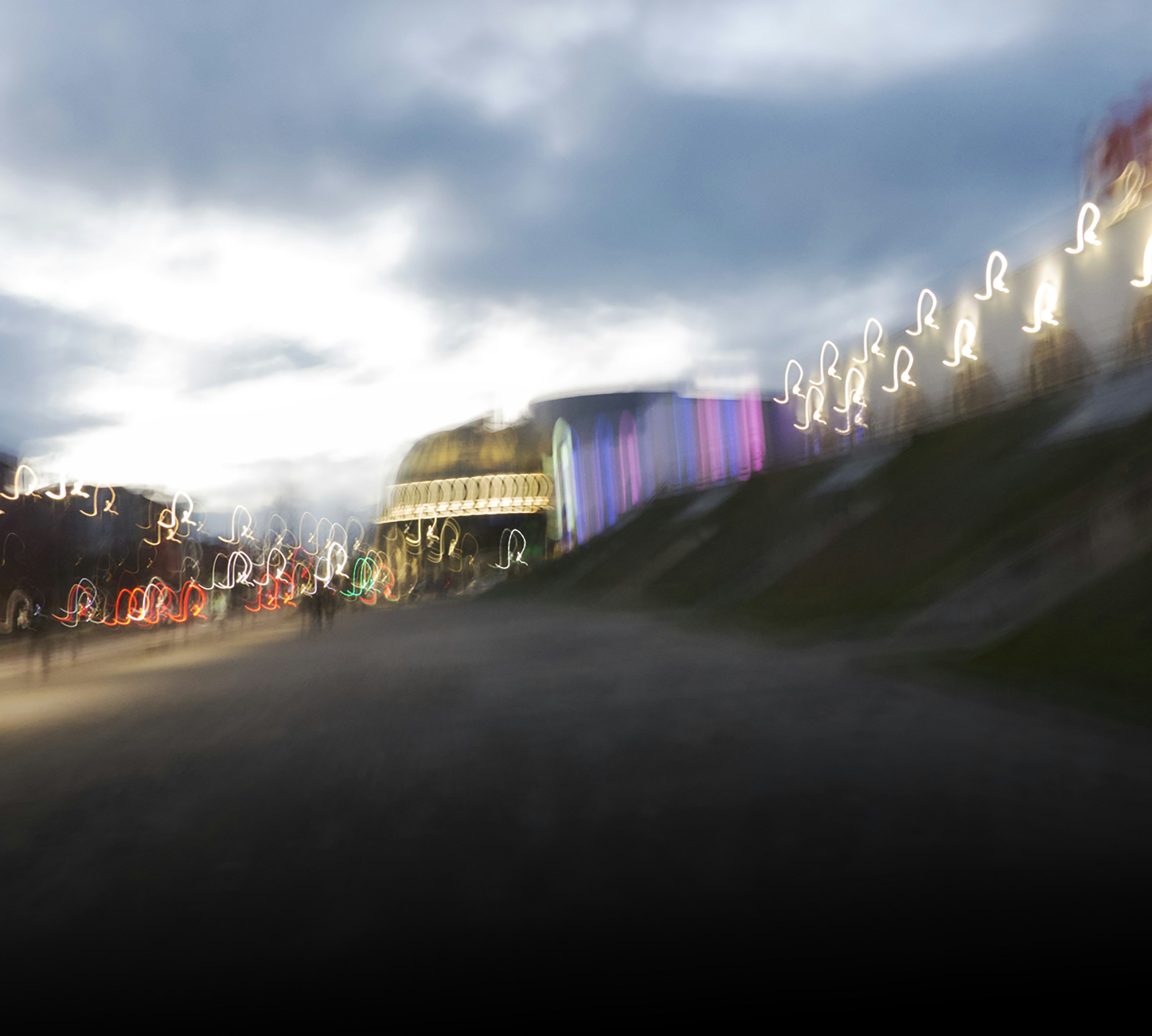 Blurred cityscape at dusk, featuring vibrant light trails from street lamps and vehicles. The scene evokes a sense of movement and energy.