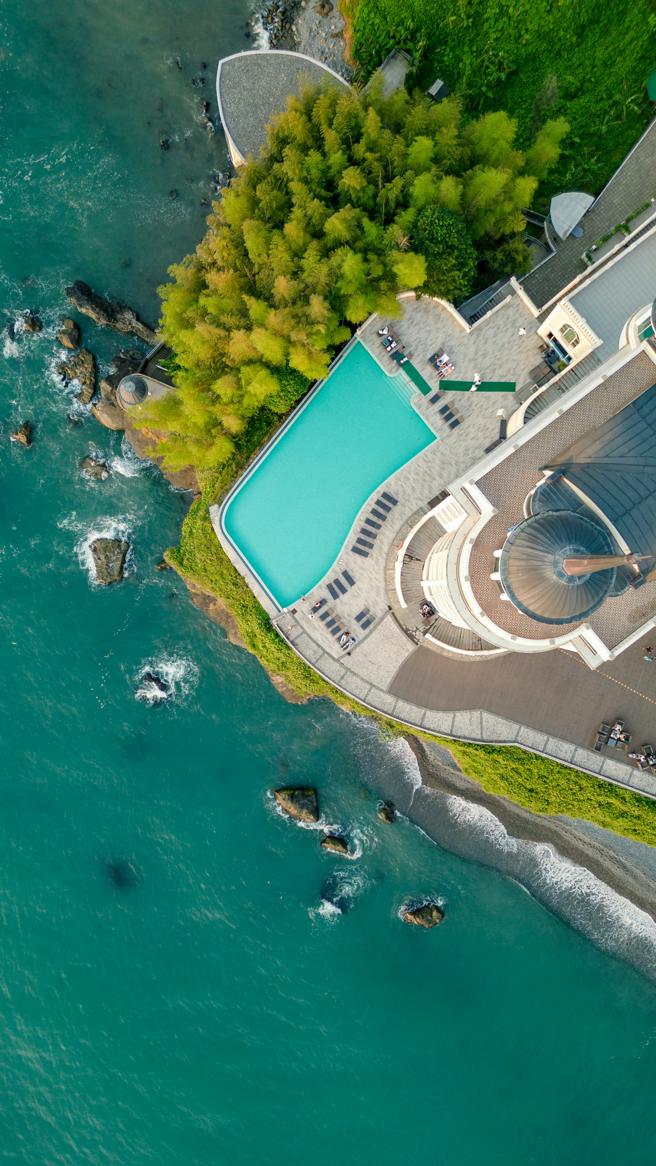 Aerial view of a coastal resort featuring a turquoise pool, lush greenery, and rocky shoreline. The harmonious blend of nature and architecture invites relaxation.