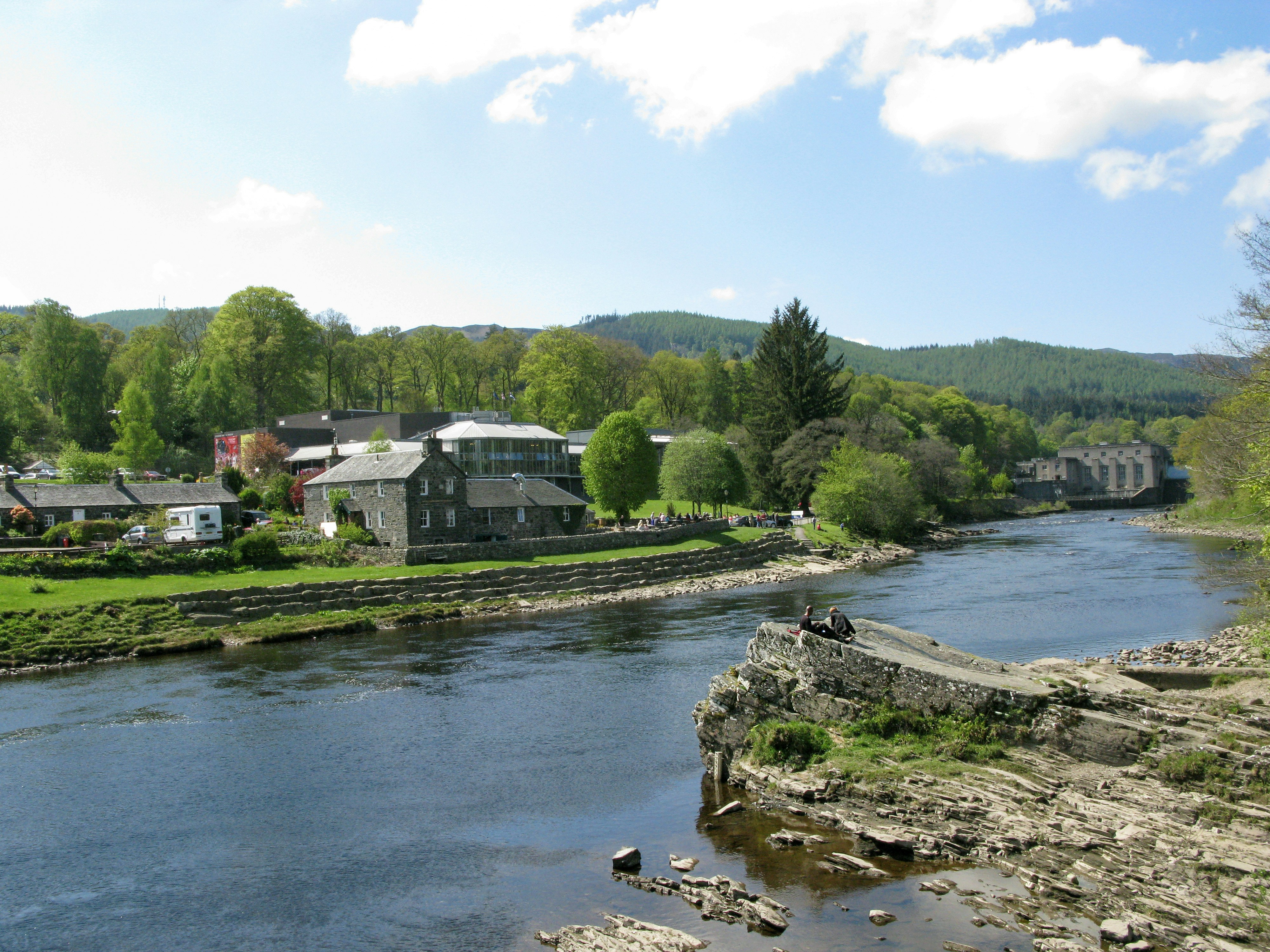 Pitlochry is a town in the Perth and Kinross council area of Scotland, lying on the River Tummel. It is historically in the county of Perthshire, | River flows through a town with green hills.