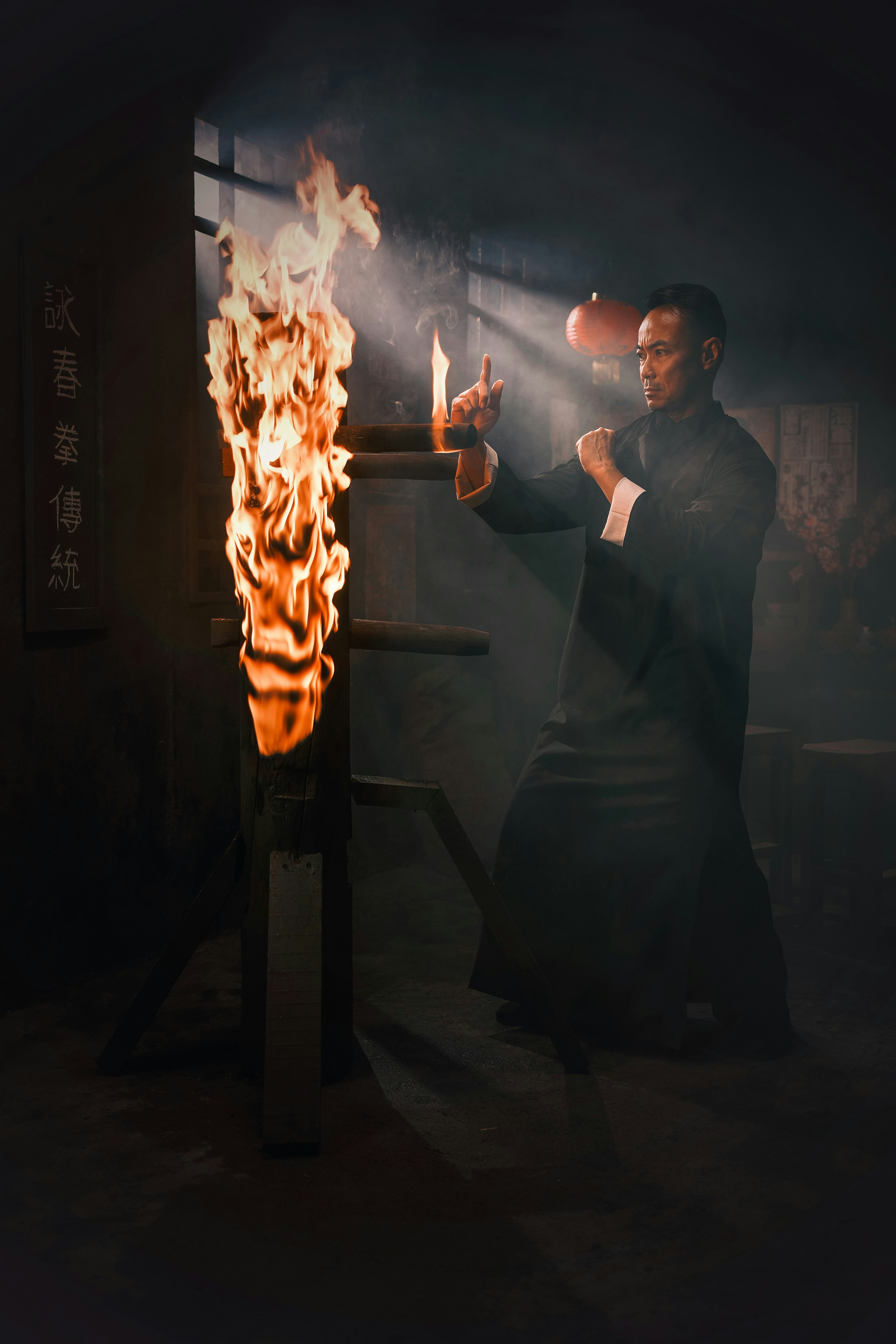 Martial artist demonstrating technique against a backdrop of flames and smoke in a traditional setting.