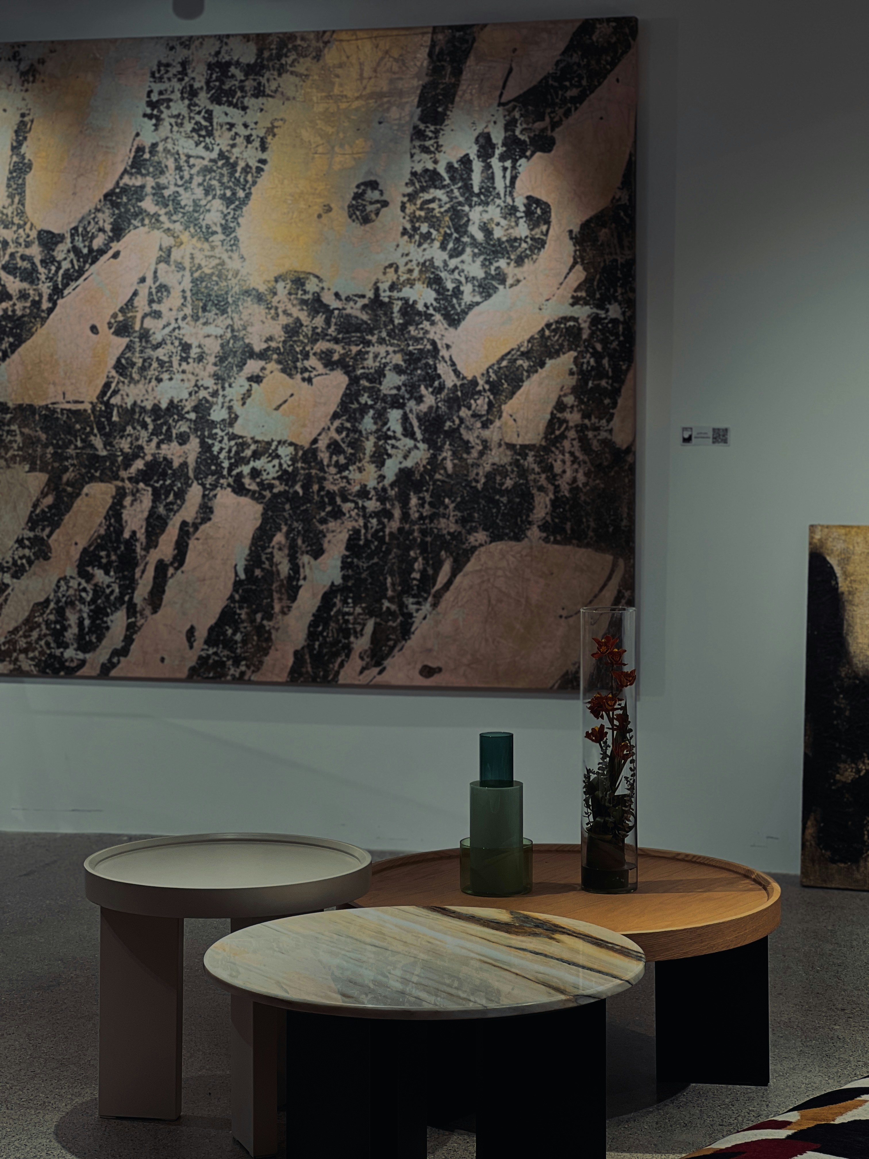 Abstract artwork dominates the wall above a trio of contemporary tables, showcasing a blend of textures and colors. A vase with flowers adds a touch of nature to the artistic setting.