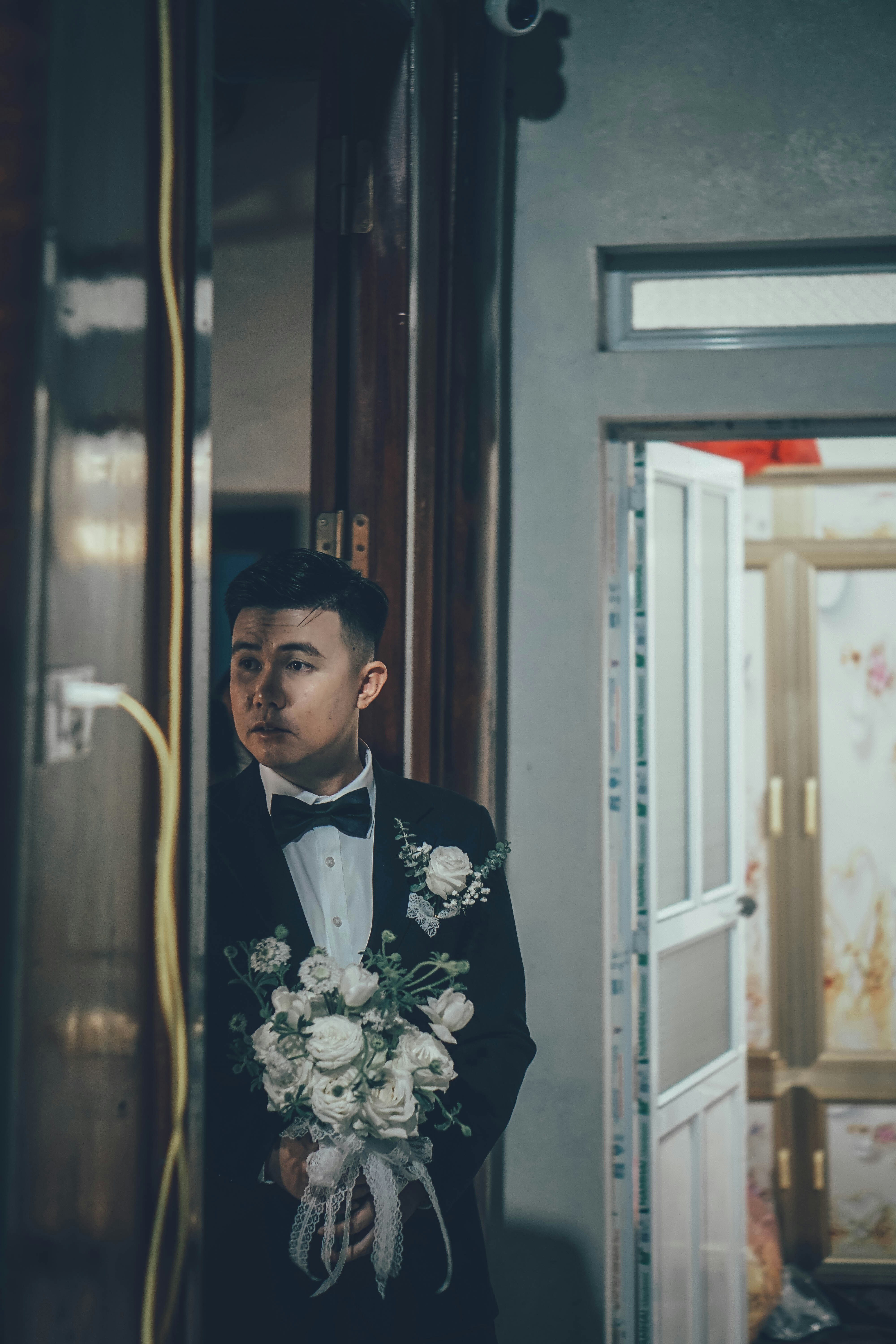 Doorway Quiet: The Groom with a White Bouquet Under Soft Indoor Light