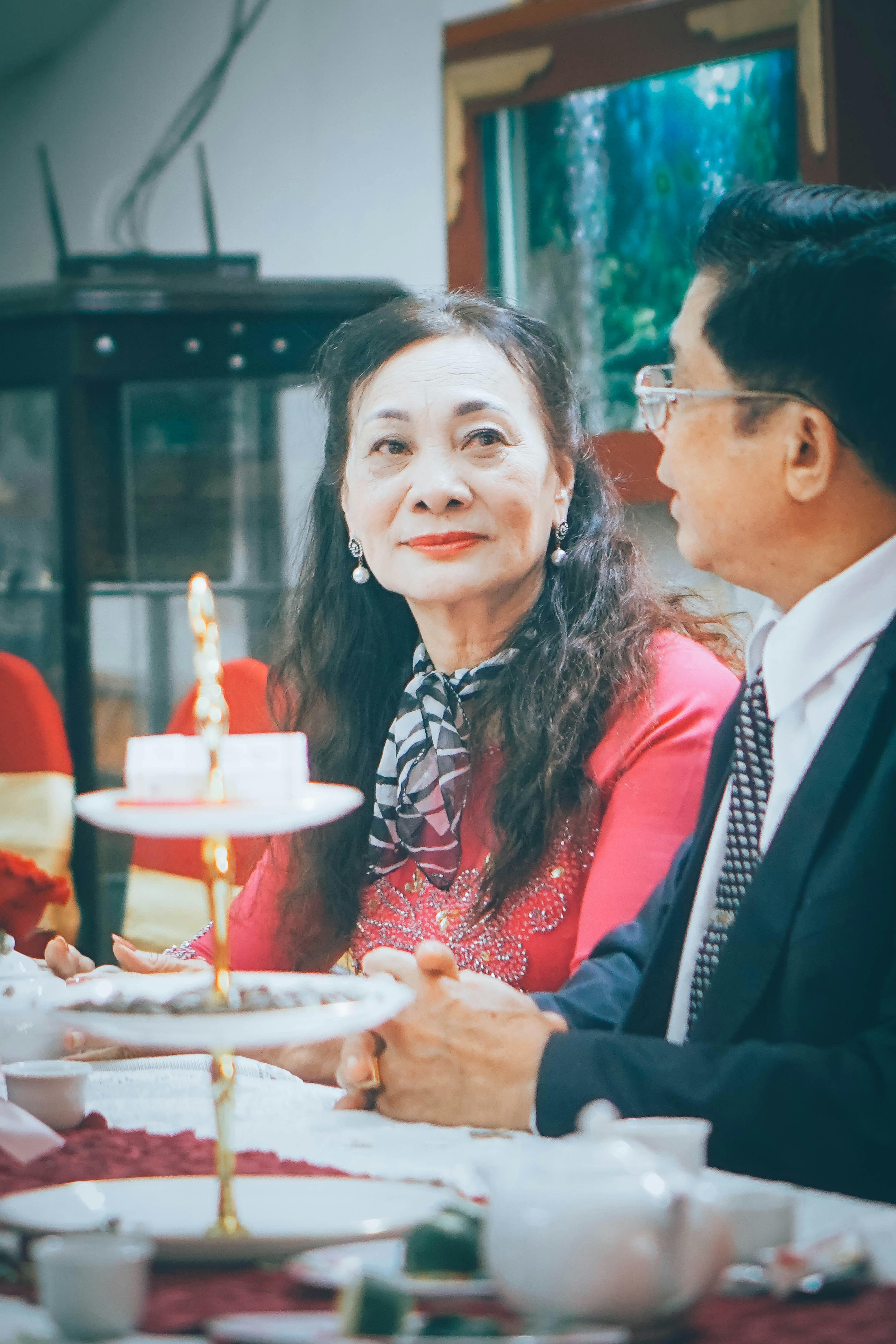 Elegant woman in a red sweater engages in conversation at a tea gathering, with a tiered serving stand in view.