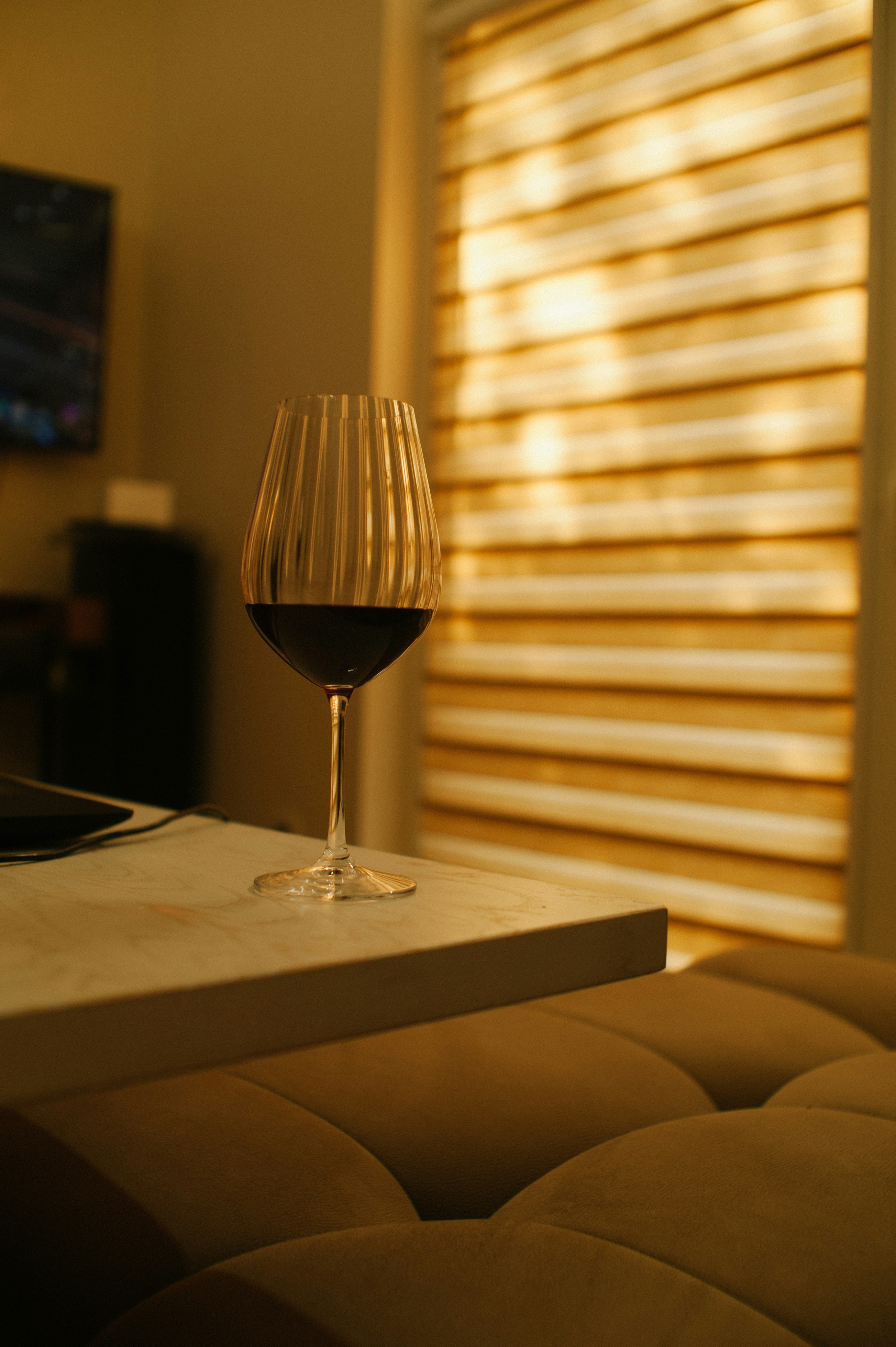 Elegant wine glass filled with red wine resting on a table, illuminated by soft golden light filtering through window shades.