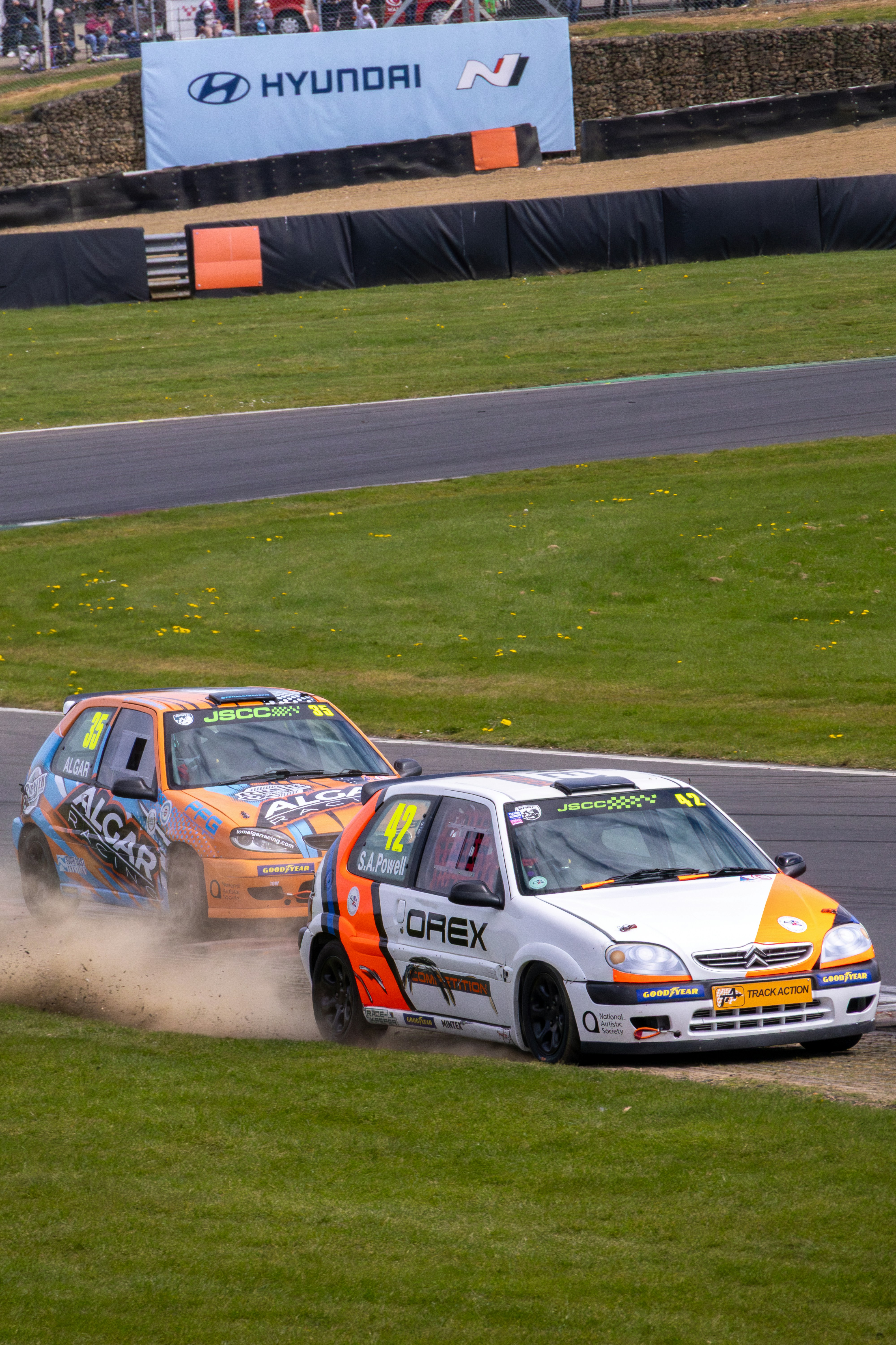 Two race cars skidding around a bend on a motorsport track, kicking up dust as they compete for position. The vibrant colors of the vehicles stand out against the green grass and asphalt.