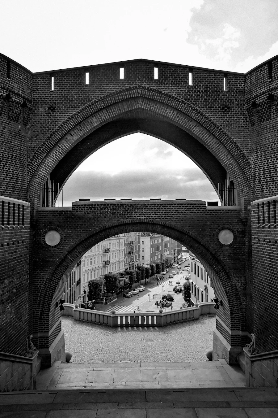 Brick archway framing a cobblestone plaza and narrow city street viewed in high-contrast black-and-white.