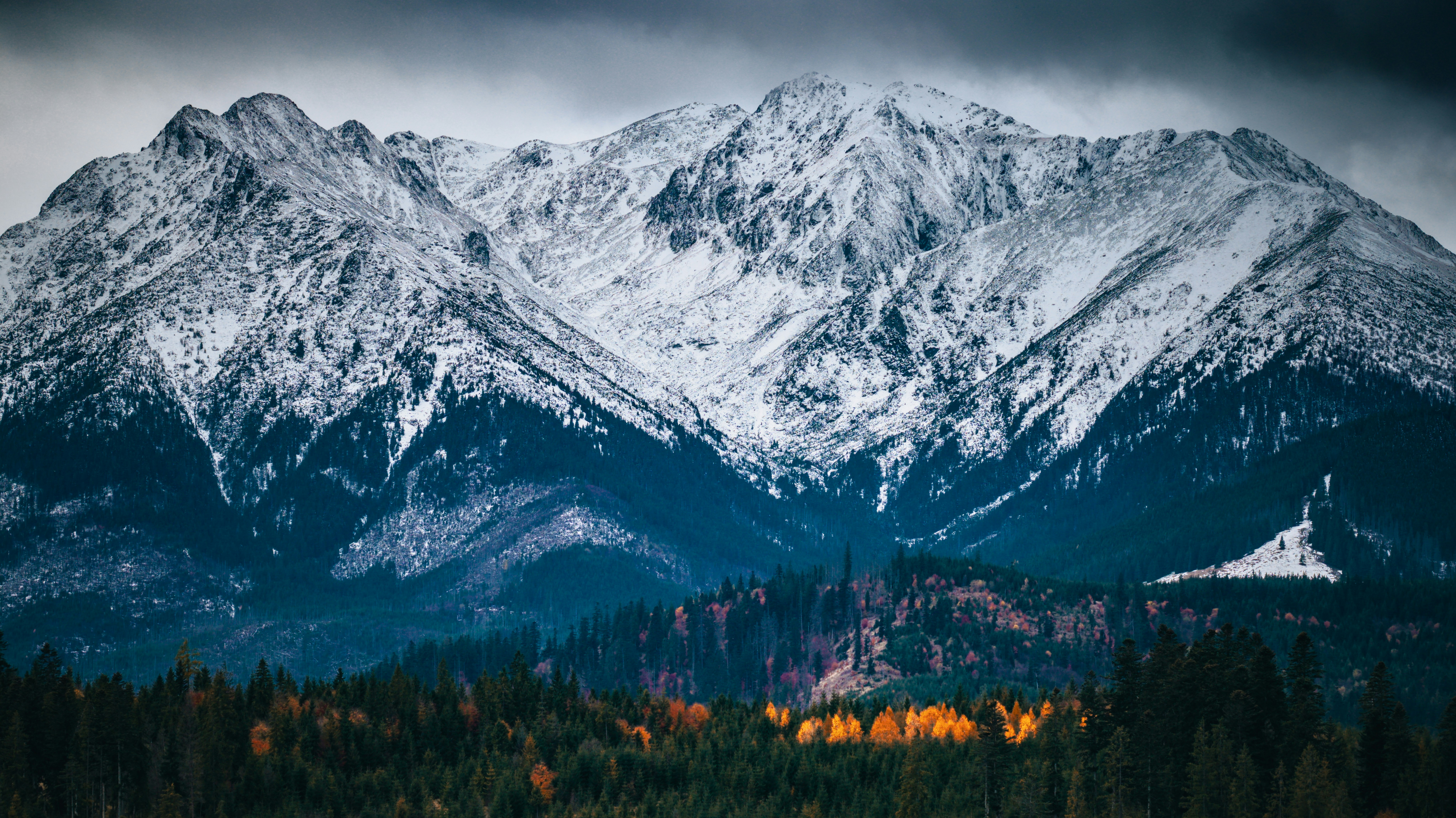 Majestic snowy peaks of the Tatra Mountains rising above dark forests and autumn colors. Captured on a cold afternoon, showing the contrast between early snow and fading fall tones. | Snow-capped mountain range with autumn forest below.
