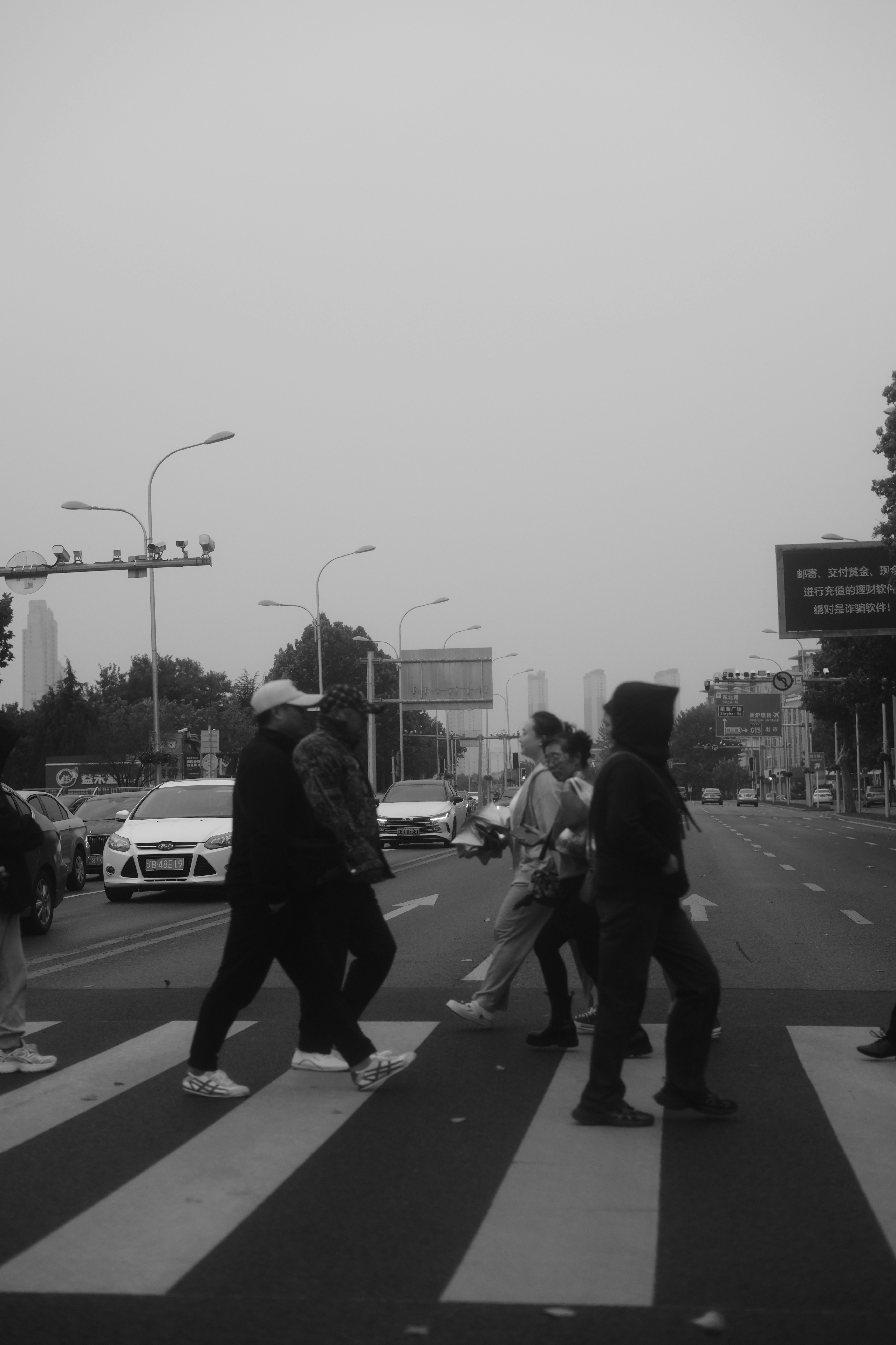 Pedestrians navigating a crosswalk on a busy city street, with vehicles waiting in the background. The monochrome palette emphasizes the dynamic urban environment.