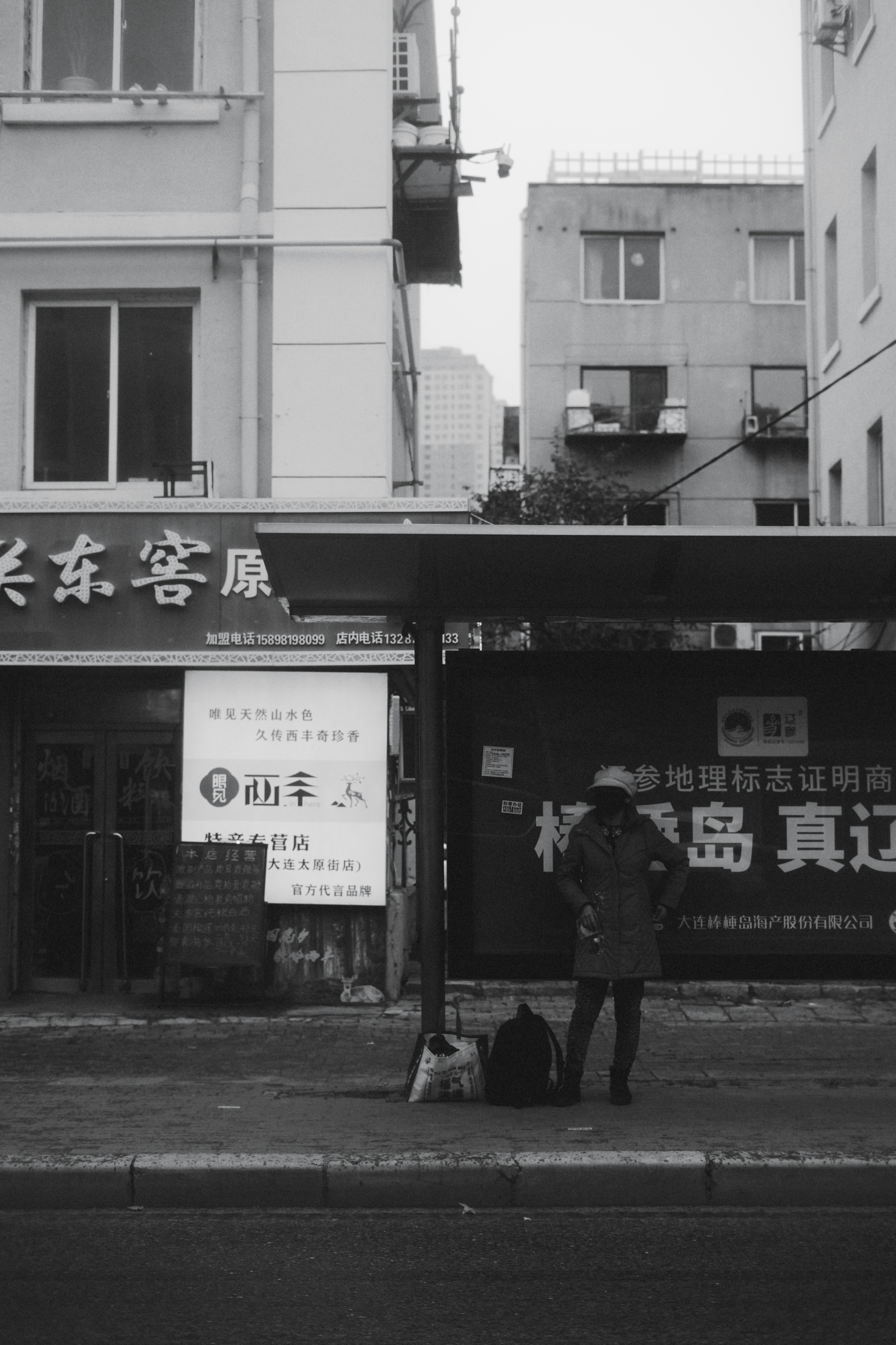 A lone figure stands at a bus stop amidst a backdrop of urban architecture, with signs in a mix of languages hinting at the locale's cultural diversity.