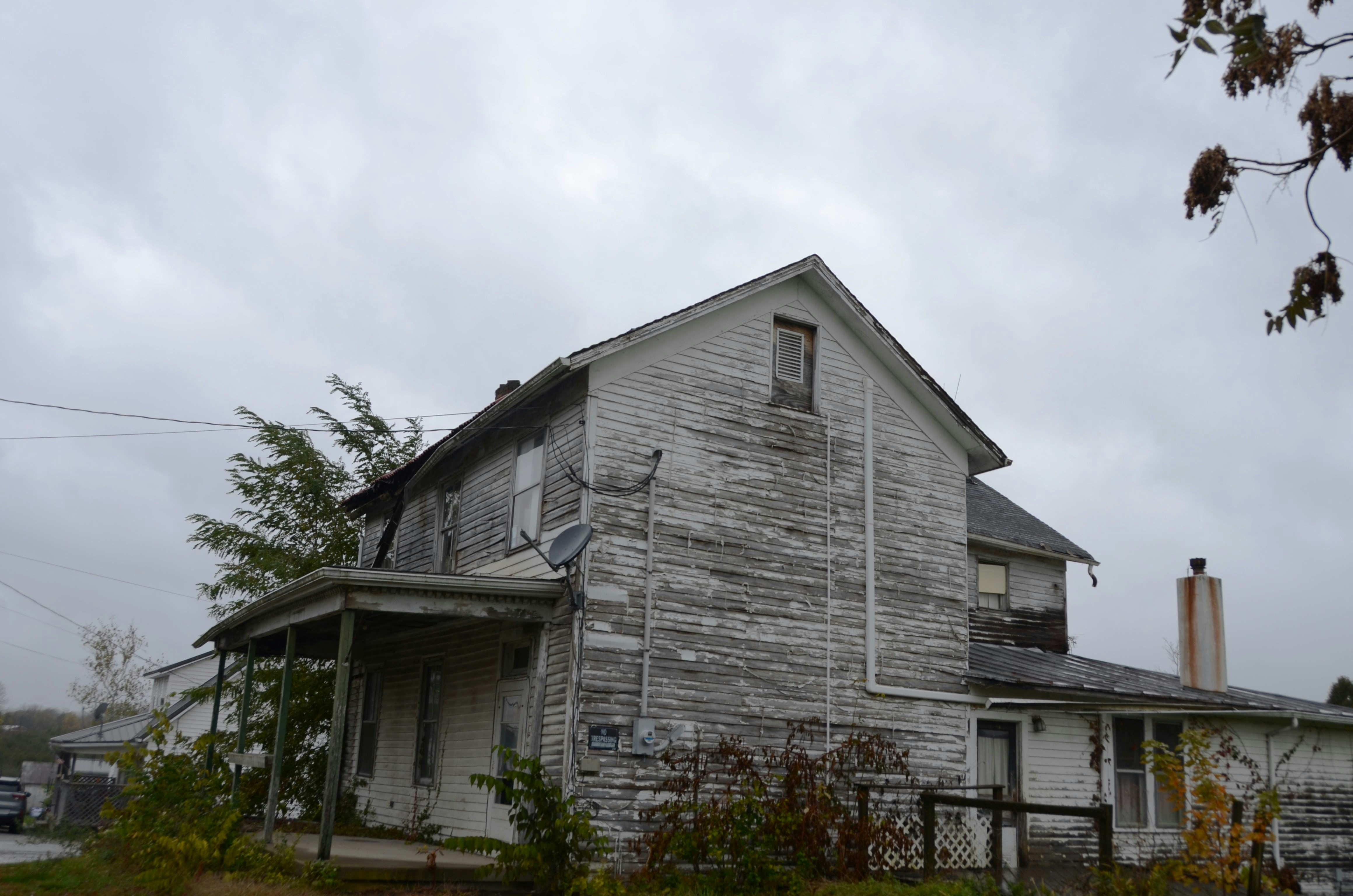 Weathered farmhouse with peeling paint and overgrown vegetation under a cloudy sky, showcasing the passage of time.