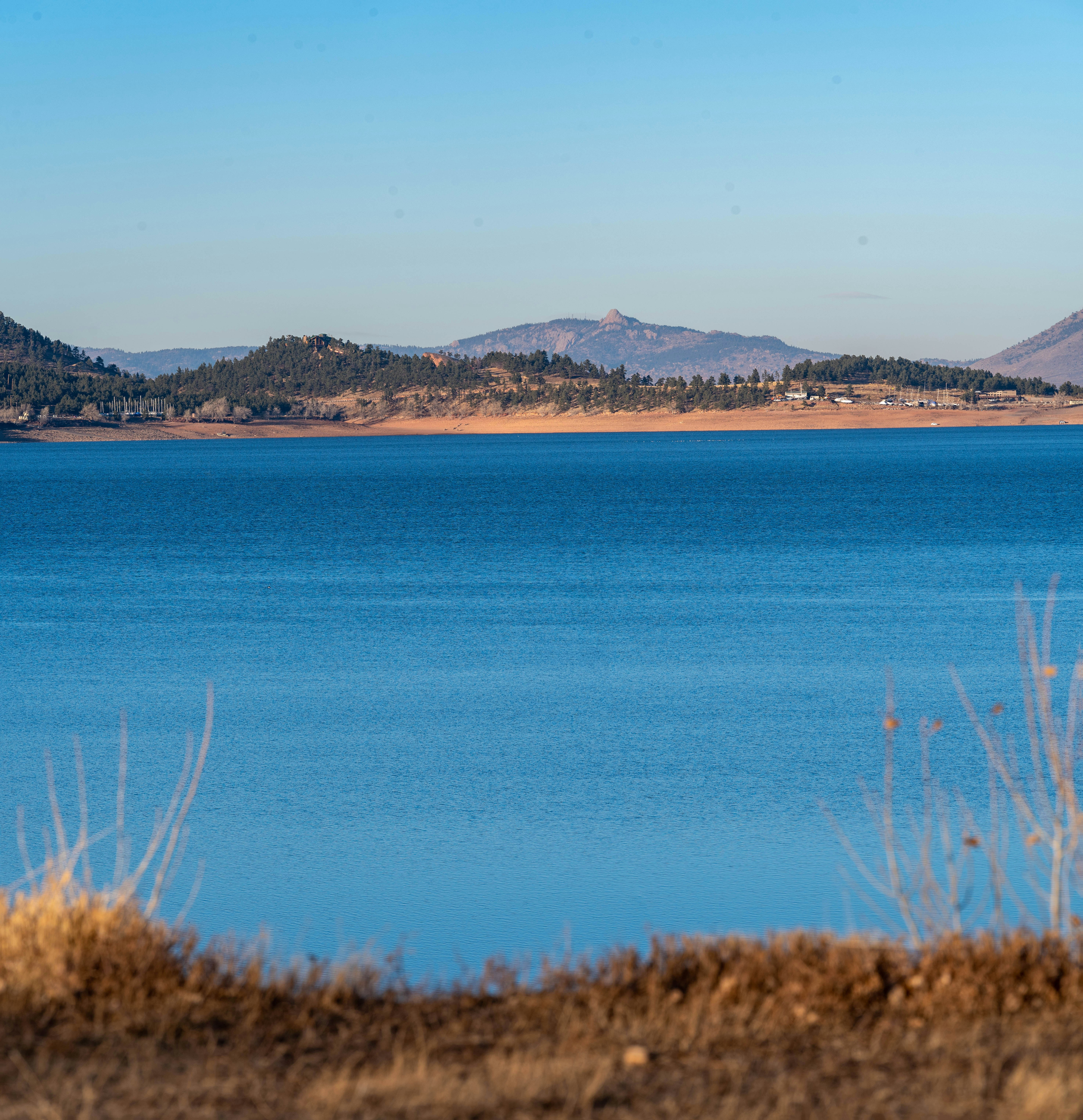 Serene lake reflecting distant mountains under a clear blue sky, with dry grass framing the foreground.