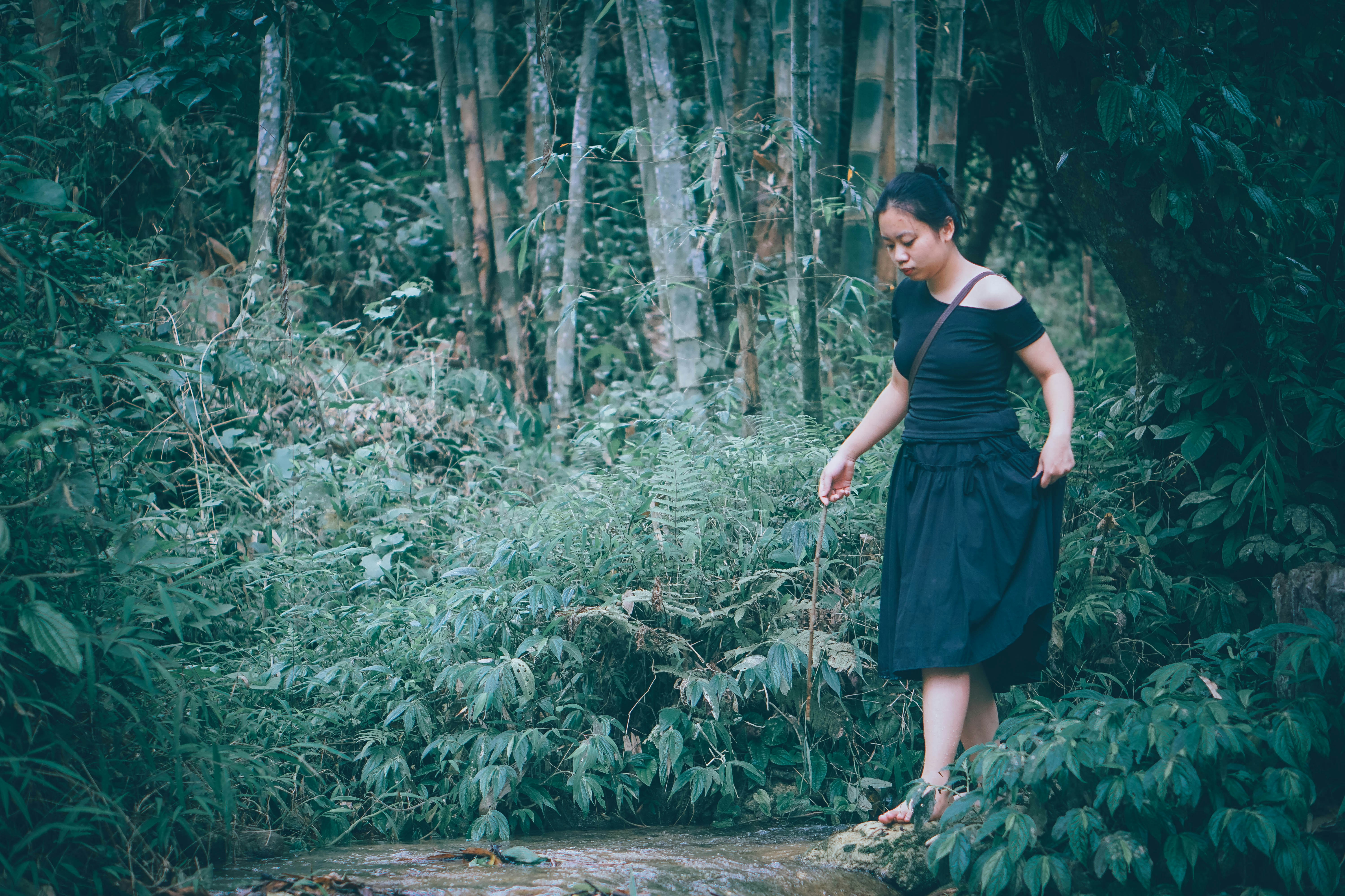 Woman in a black dress carefully steps over a stream surrounded by dense foliage and bamboo. The scene evokes tranquility and connection with nature.
