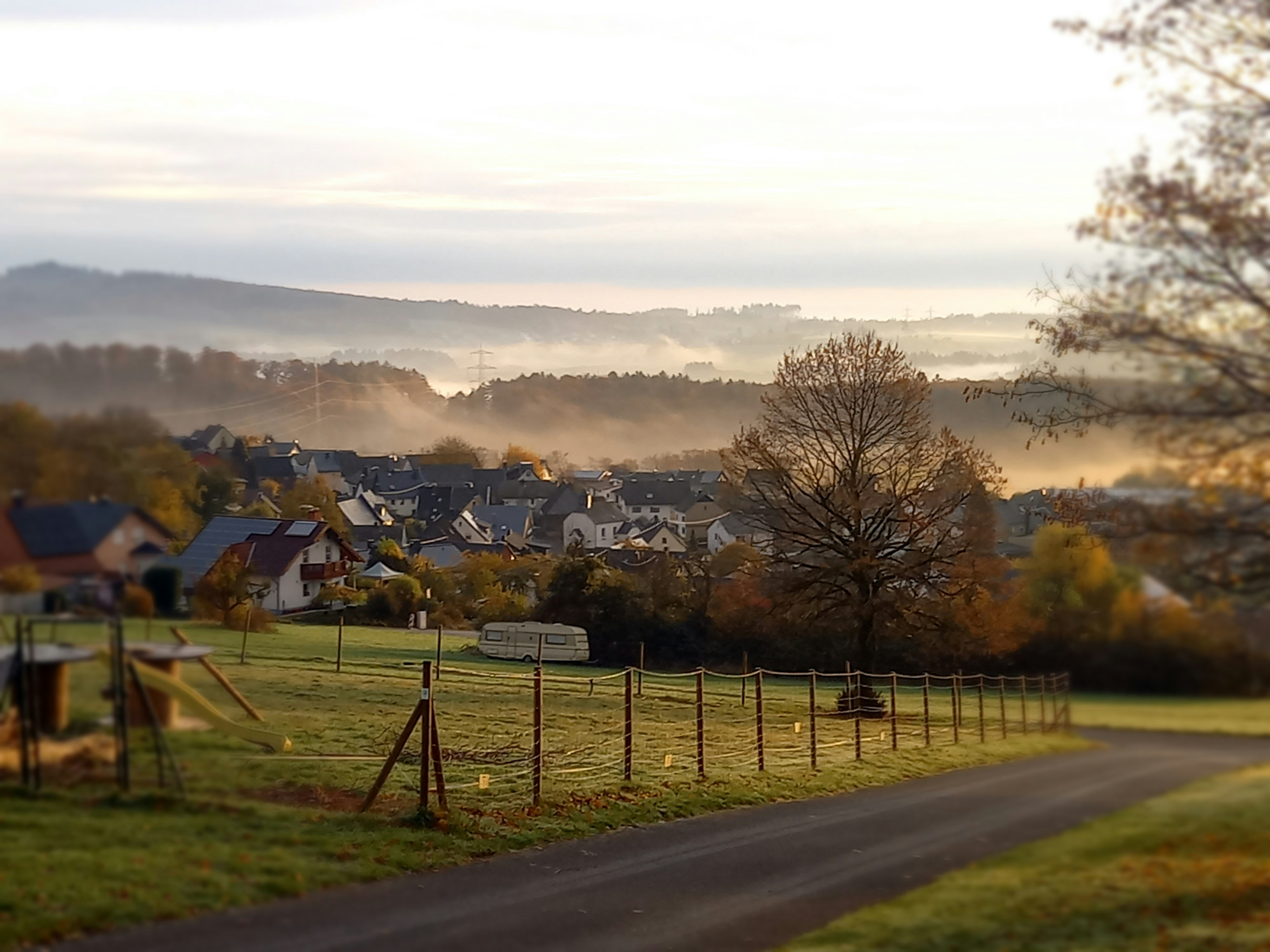 A tranquil village landscape at dawn, enveloped in mist, with houses nestled among trees and rolling hills. The scene captures the peaceful essence of early morning light.