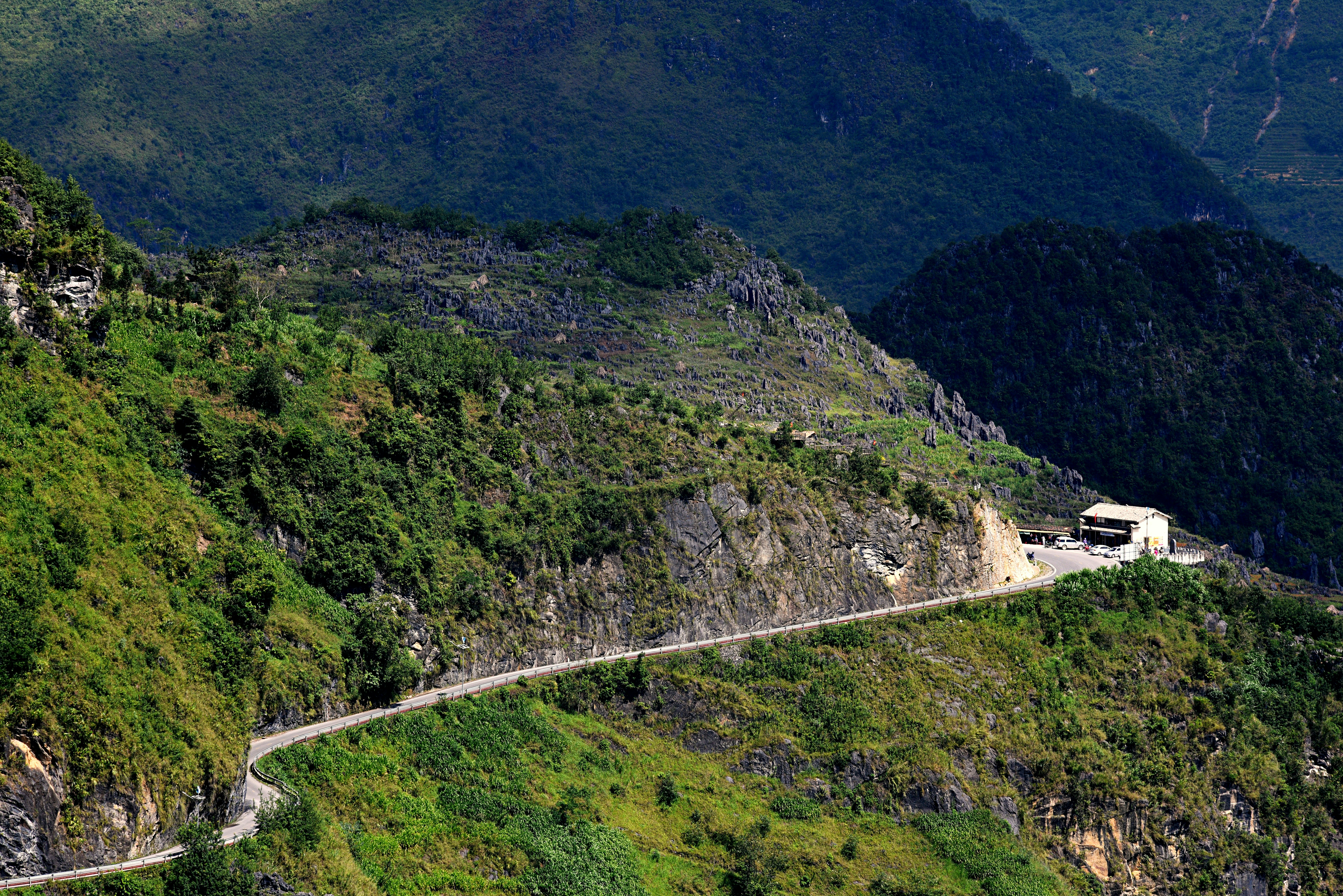 A winding road meanders through lush green hills, leading to a distant structure nestled among rocky terrain.