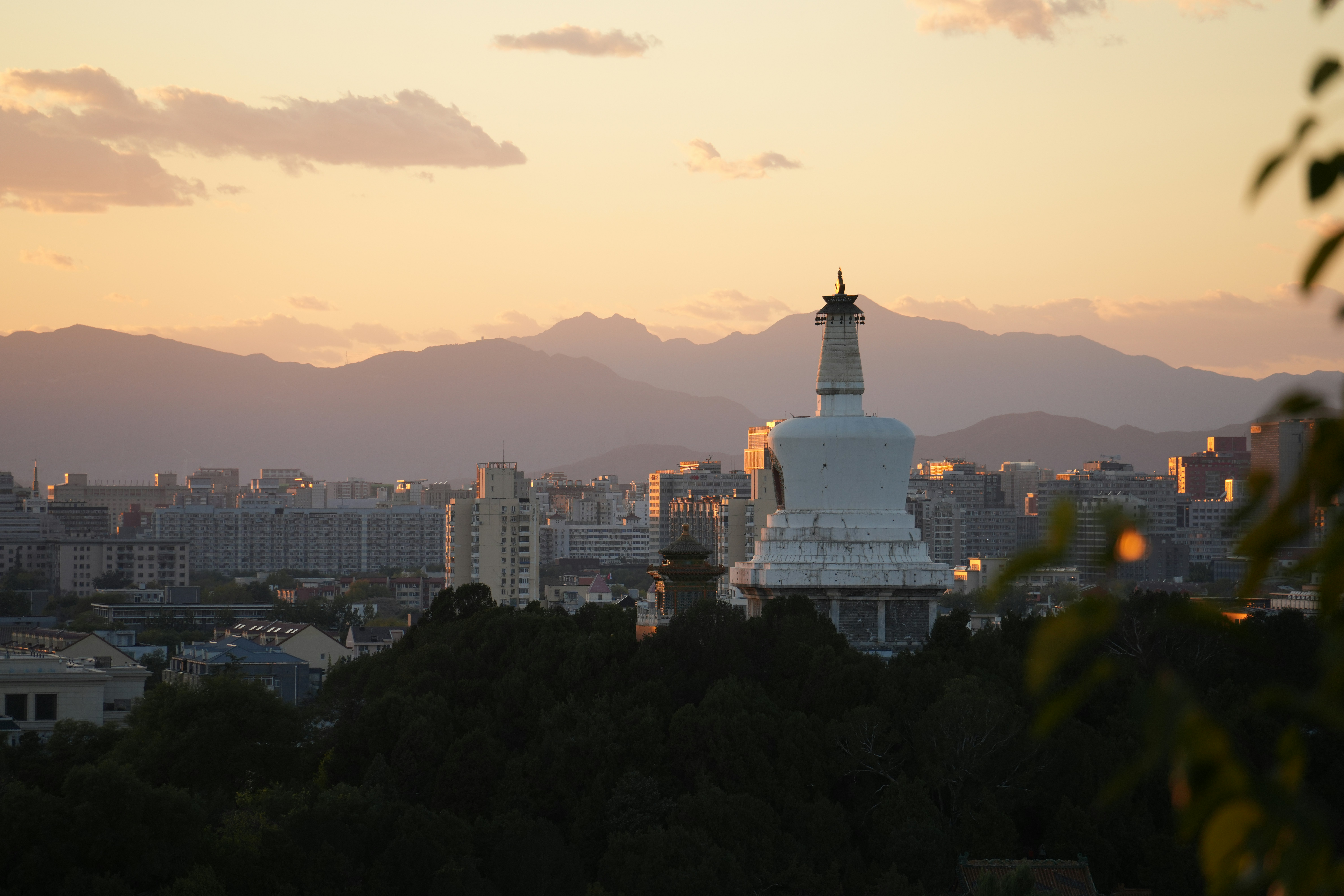 Historic white pagoda silhouetted against a vibrant sunset, surrounded by urban landscape and distant mountains.