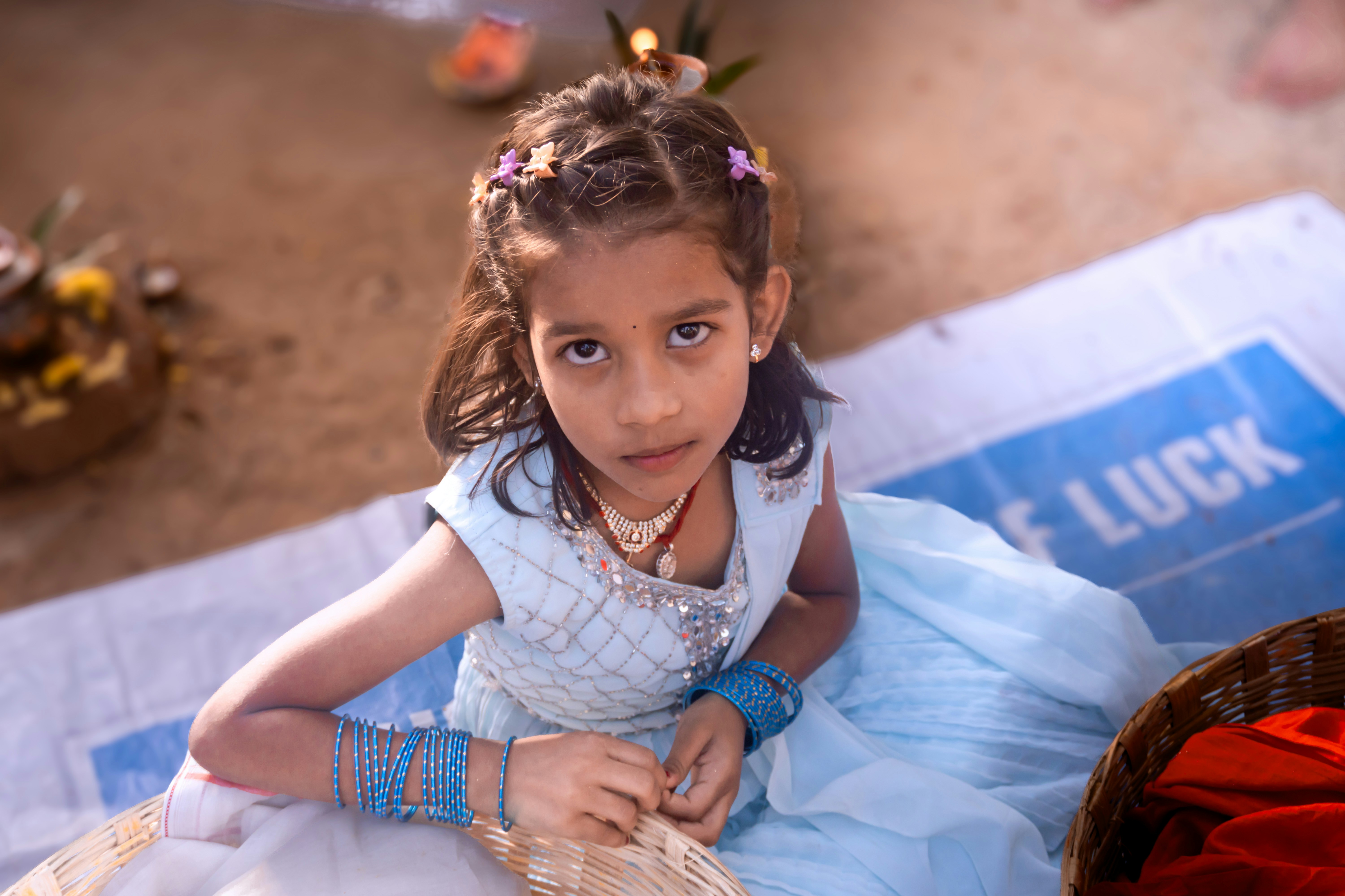 A young girl dressed in a light blue traditional outfit gazes thoughtfully, surrounded by festive decorations and a woven basket. Her expression reflects a moment of contemplation amidst celebration.